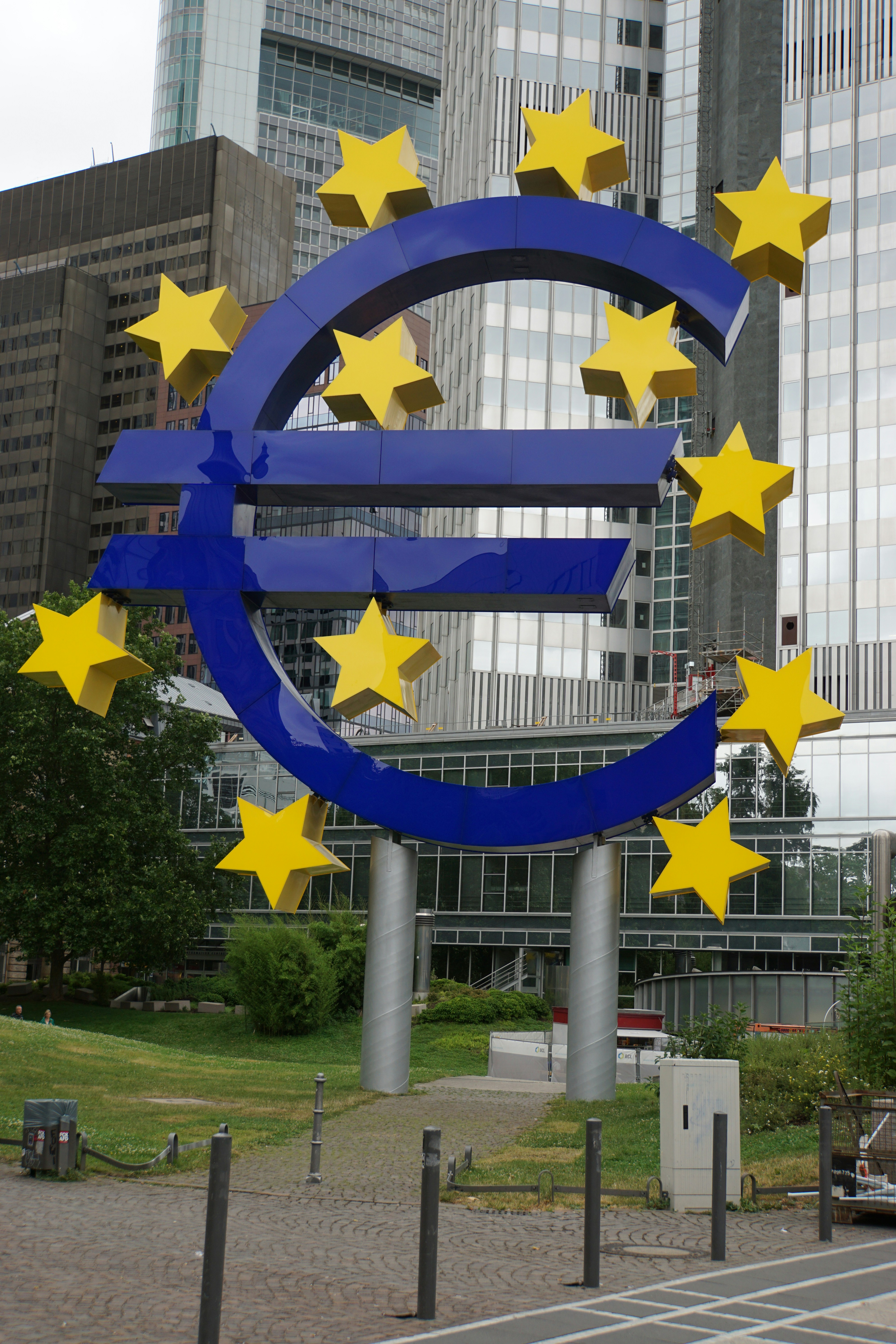 Euro sign sculpture: blue arc and yellow stars rising in front of modern towers