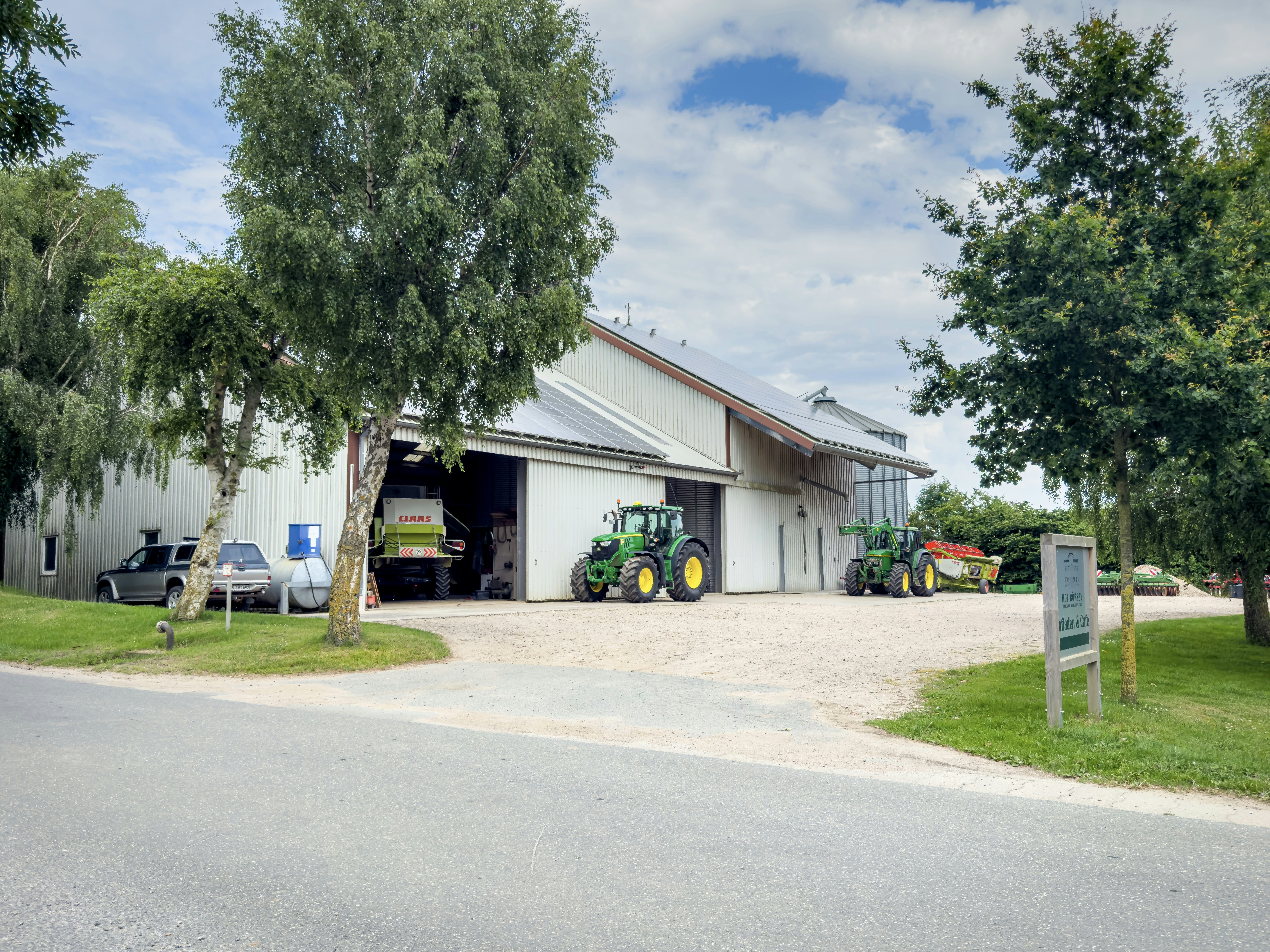 Farm machinery sits outside a metal shed with a gravel yard and surrounding trees, suggesting a rural equipment facility.
