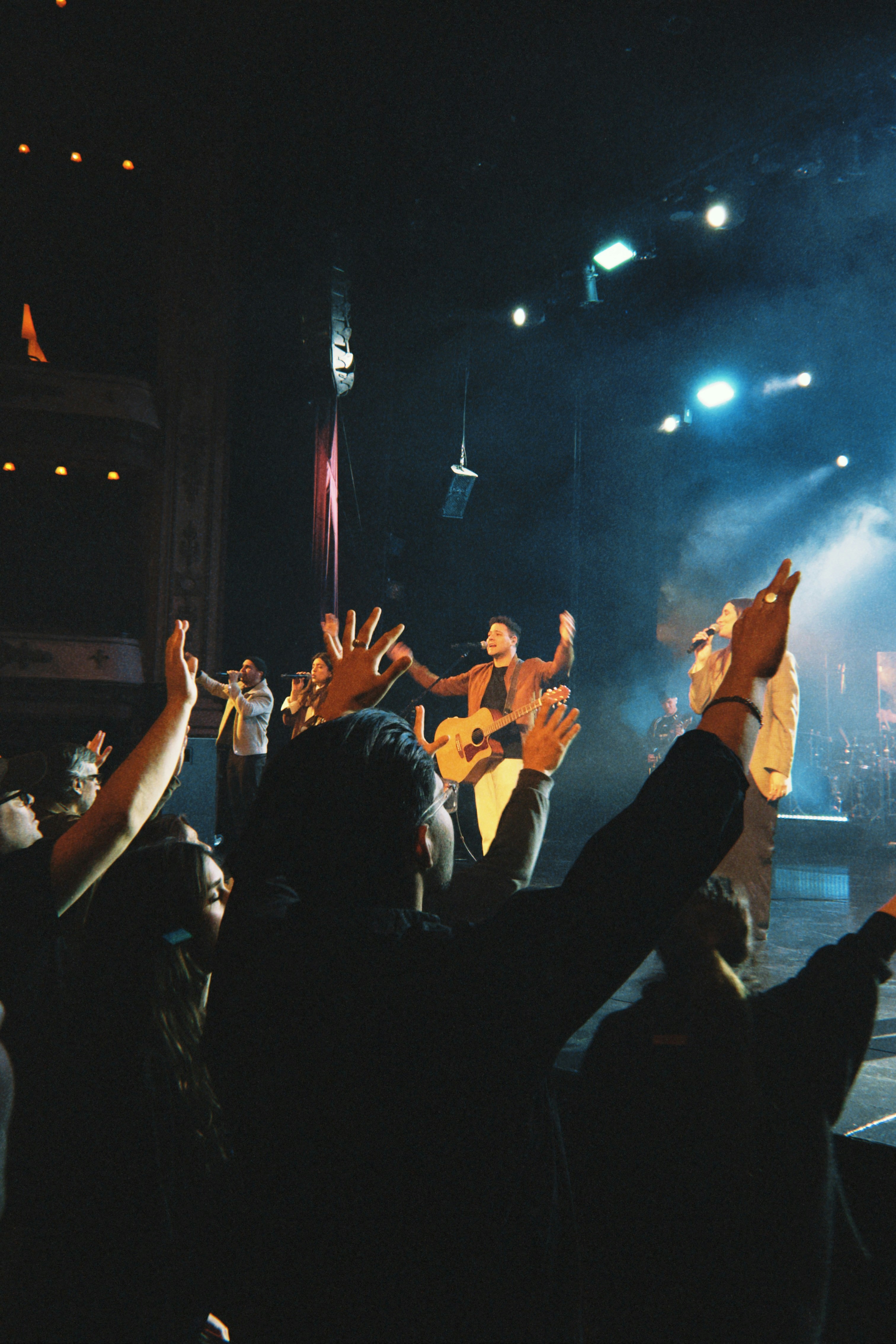 Energetic crowd with raised hands facing a lit concert stage; a guitarist and vocalists perform under vibrant stage lighting, capturing a live music moment in a theater venue.