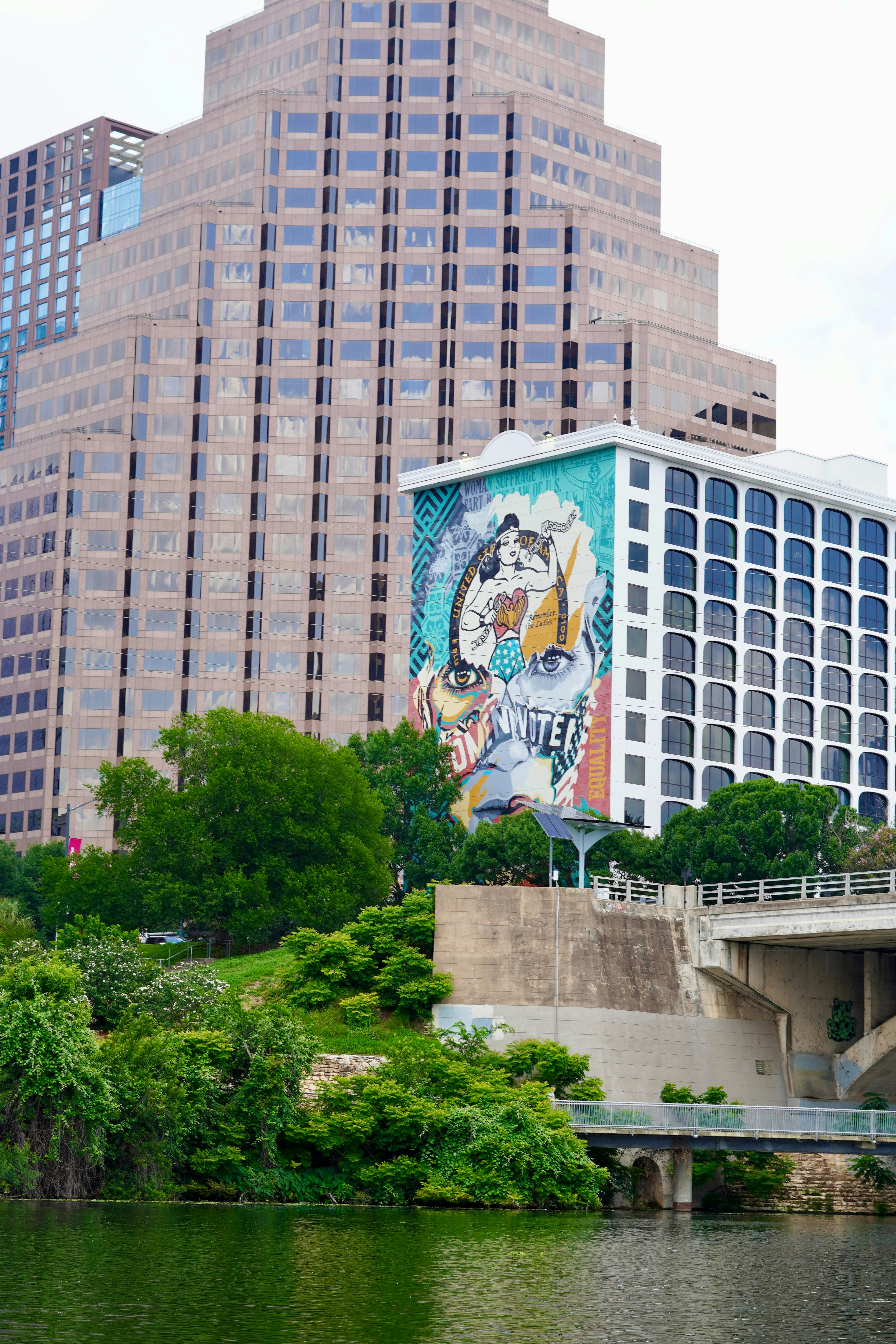 Vibrant mural-adorned building along a river with greenery and a concrete bridge in the foreground.