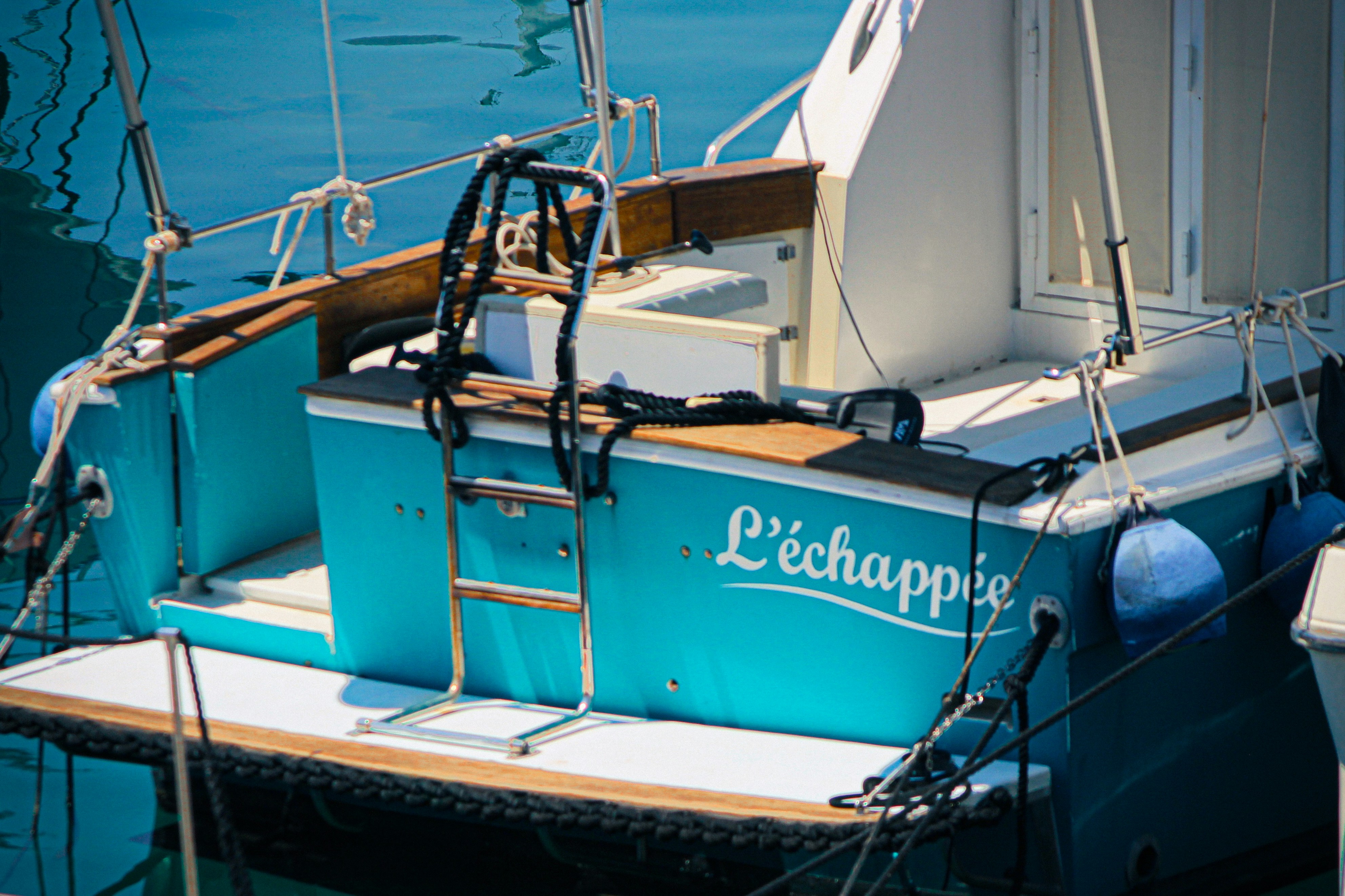 Bright turquoise aft of a docked sailboat with white rigging and wooden accents; the vessel’s name L’échappée visible along the side.