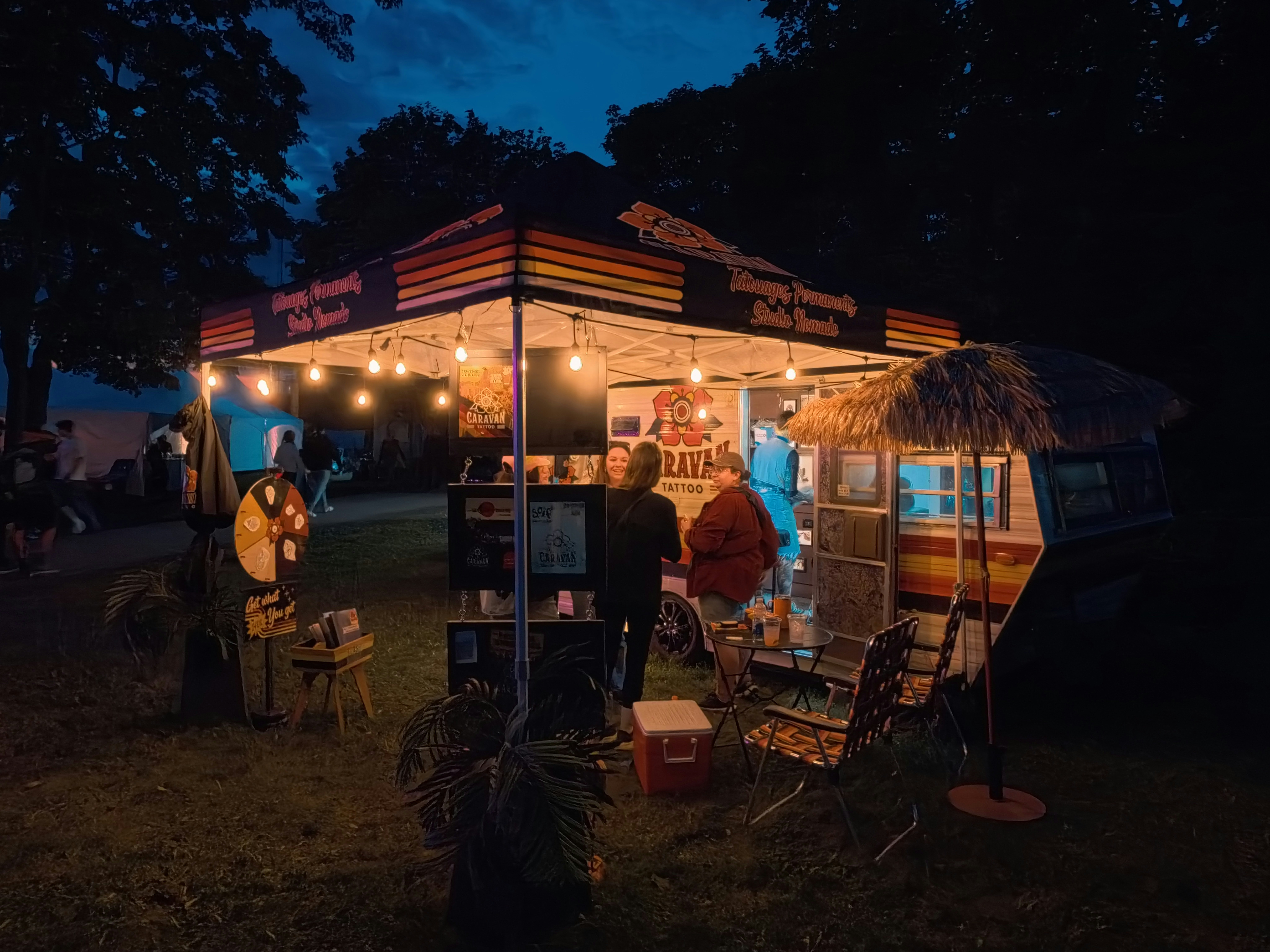 Warm-lit stall at a night market with people gathered around a colorful booth and a thatched umbrella; a framed setup and seating create a welcoming, informal scene.