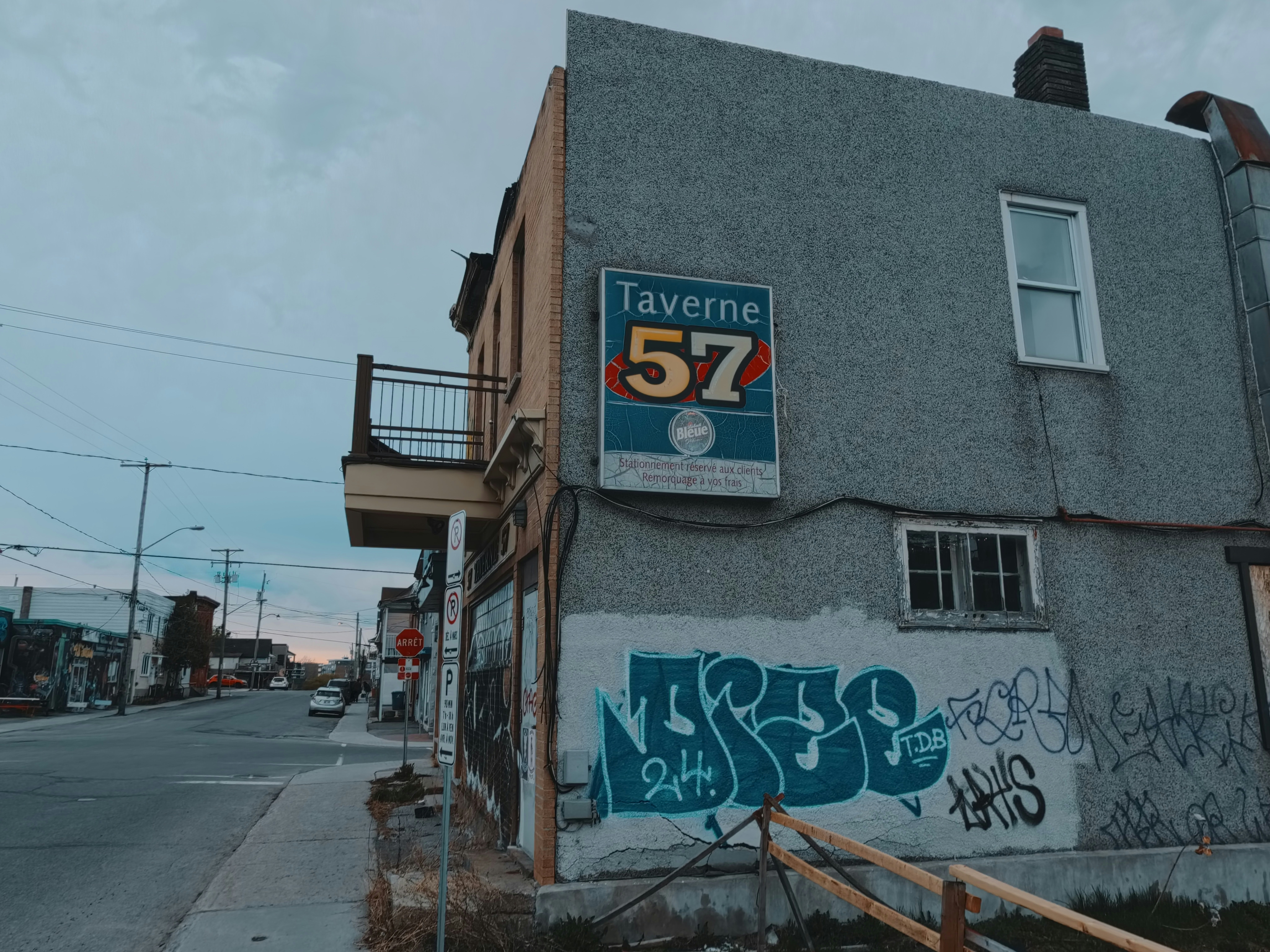 Graffiti-sprayed corner of a weathered building with a Taverne 57 sign, captured along a quiet street at dusk. The scene emphasizes urban textures and street-art.