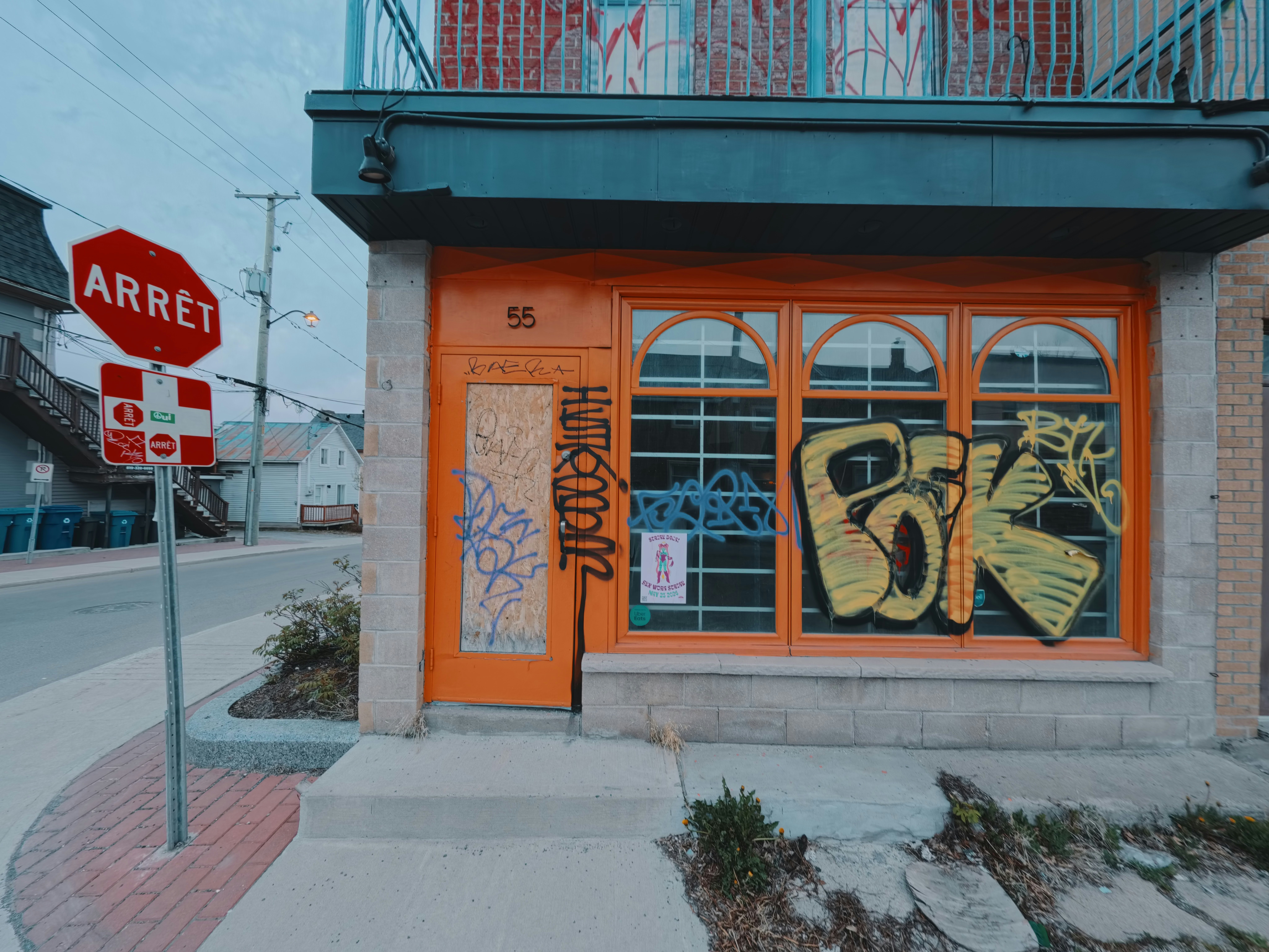 Bright orange storefront with large graffiti windows along a quiet street; stop sign and sidewalk framing the scene.