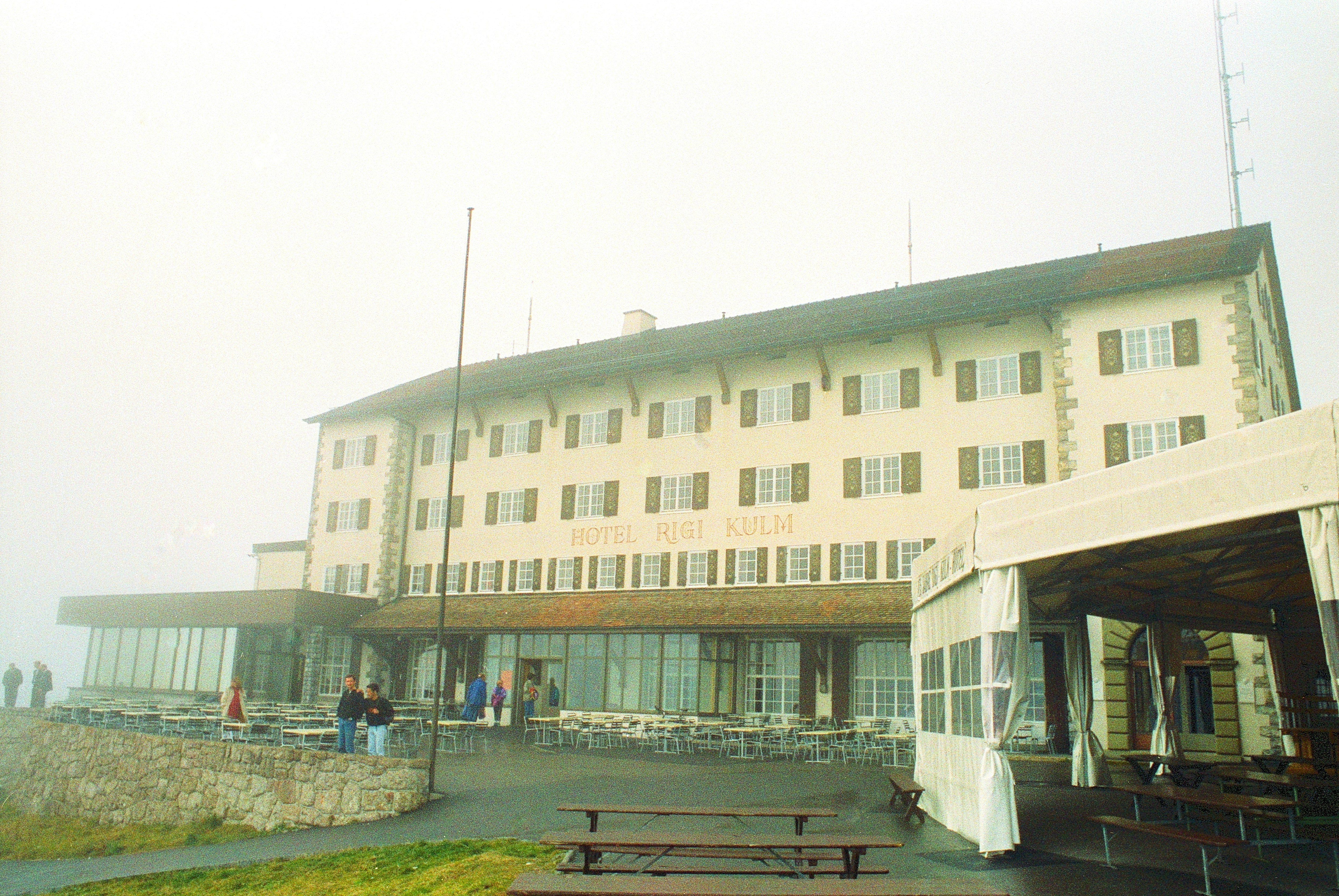 A misty early 1990s color film photograph of the iconic Hotel Rigi Kulm at the summit of Mount Rigi in central Switzerland. The large traditional Swiss hotel with its dark shuttered windows emerges dramatically from thick fog, its name clearly displayed across the facade. Empty outdoor terrace tables and benches await visitors, while a handful of tourists in warm jackets mill about despite the grey, cloud-shrouded conditions. A white marquee canopy frames the right side, adding to the quiet, off-season alpine atmosphere.