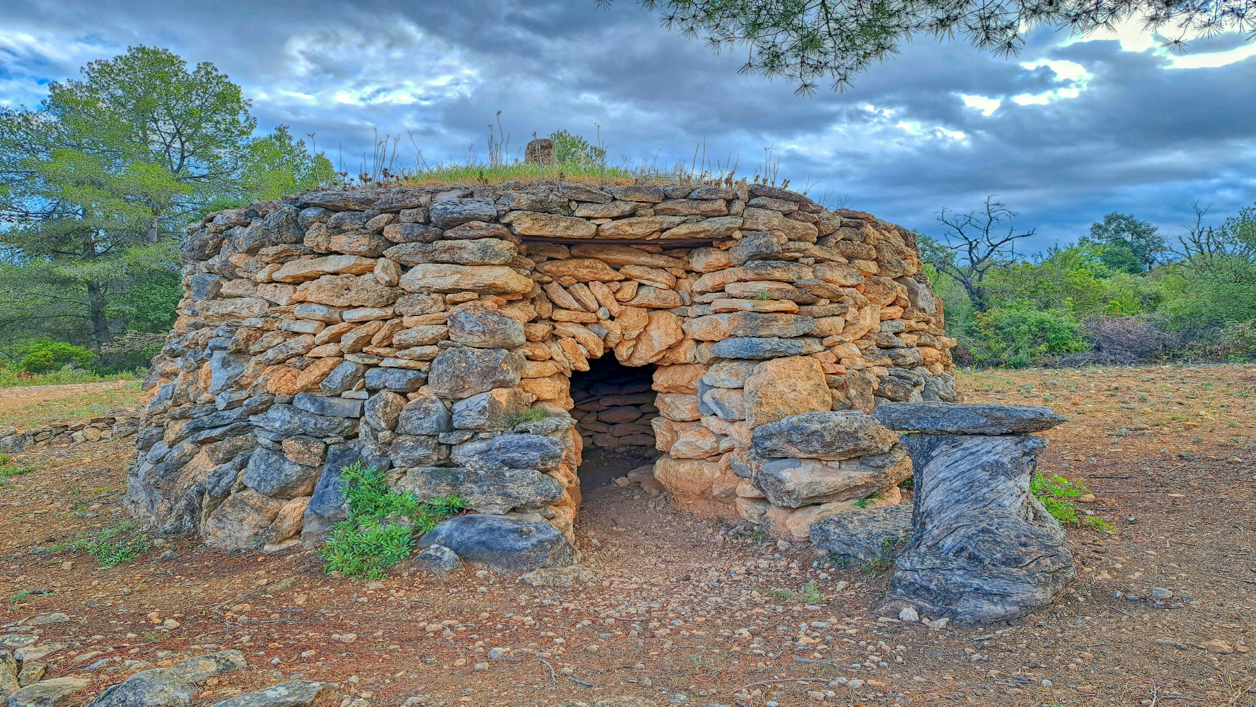 Circular stone hut with a dark doorway sits in a dry, earthy clearing; a stone bench-like structure stands to the right, under a cloudy sky.