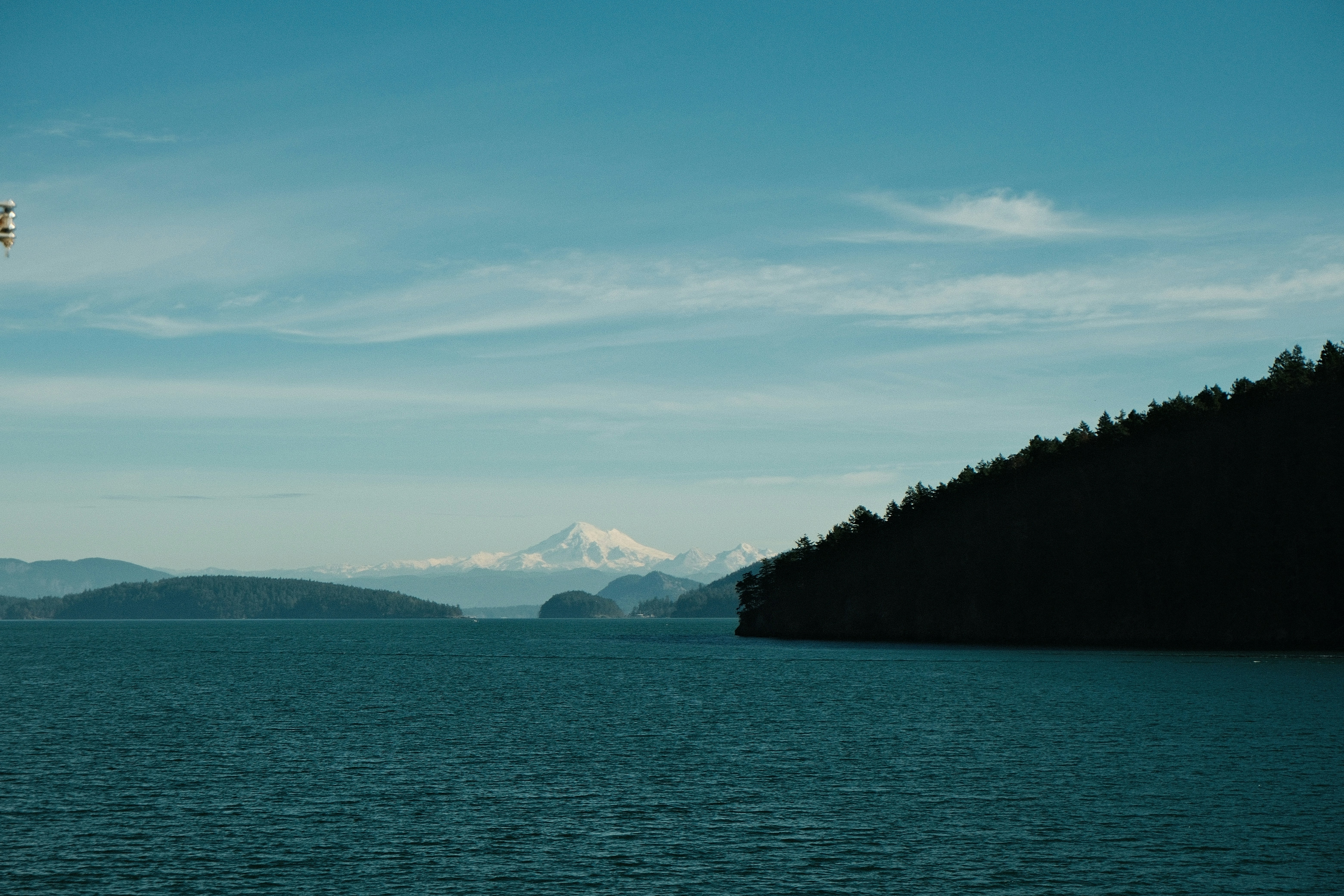 Snow-capped peak on the distant horizon reflected over calm blue water; silhouettes of shorelines frame a tranquil seascape, with a clear sky above. This is a landscape photograph.