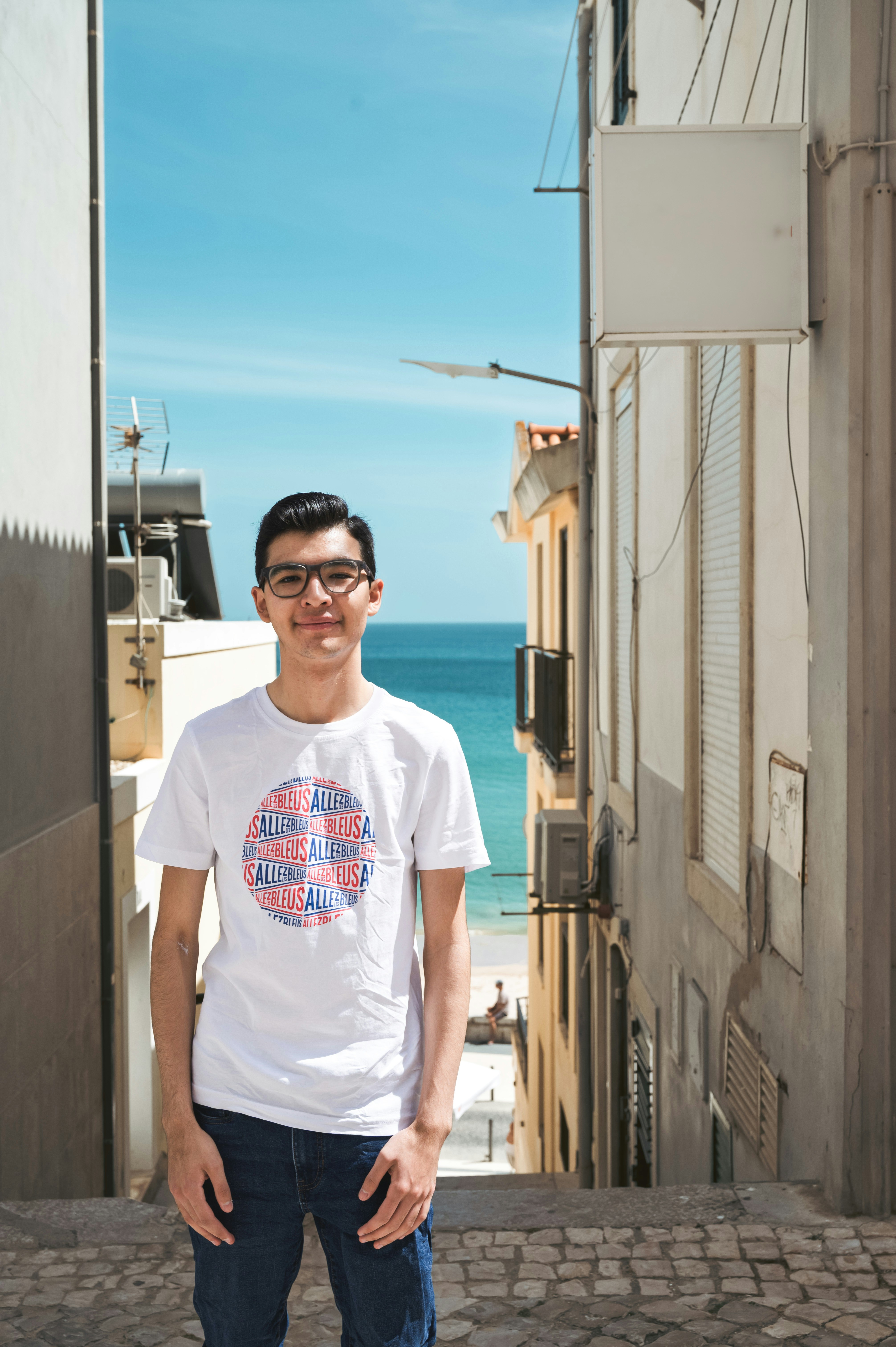 Portrait of a young man in a white graphic tee standing in a sunlit alley with the sea visible beyond. A candid urban-vacation vibe between narrow buildings and bright blue sky.