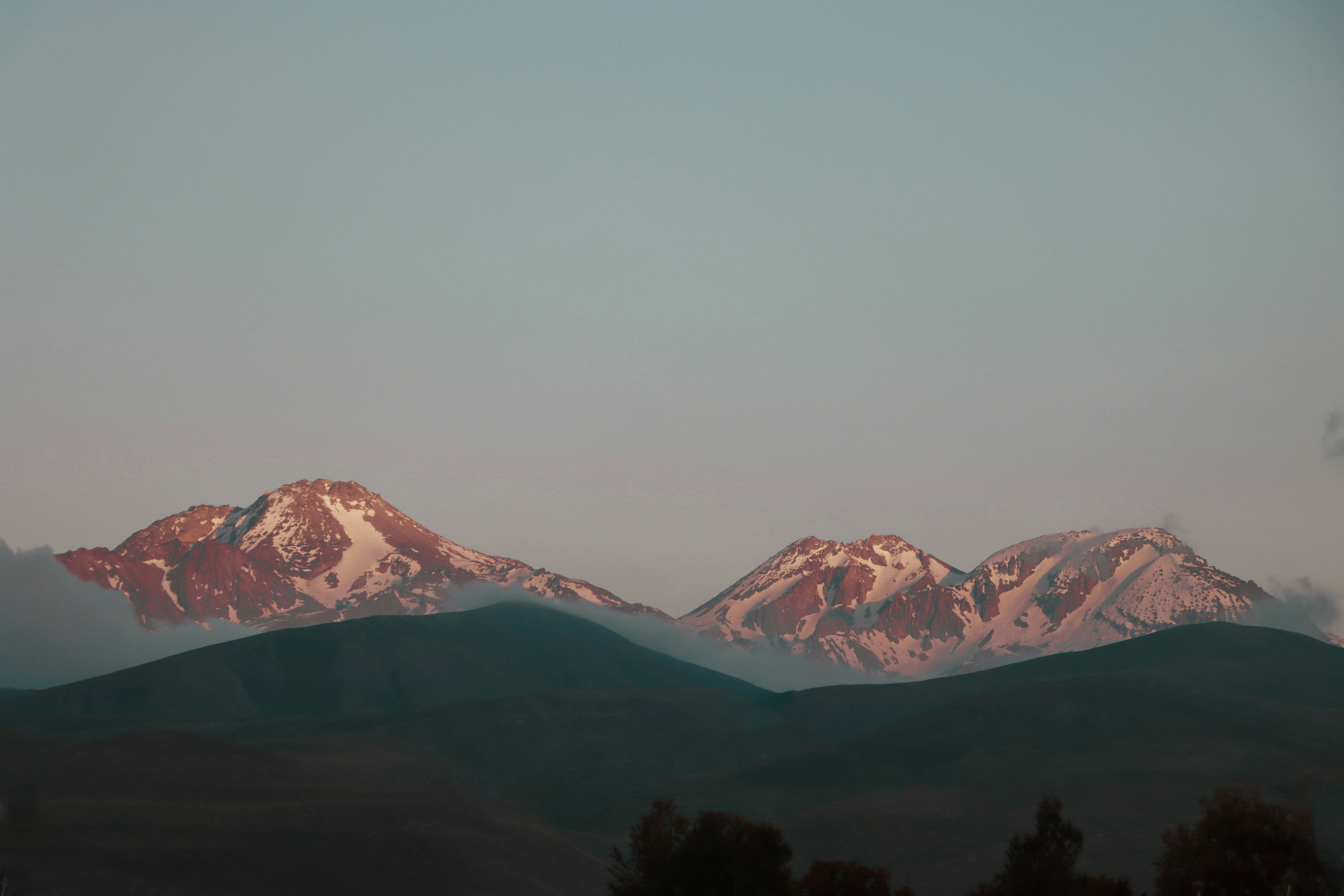 Distant snow-capped mountains rise above rolling dark hills at twilight, with a soft gradient sky and a calm foreground.