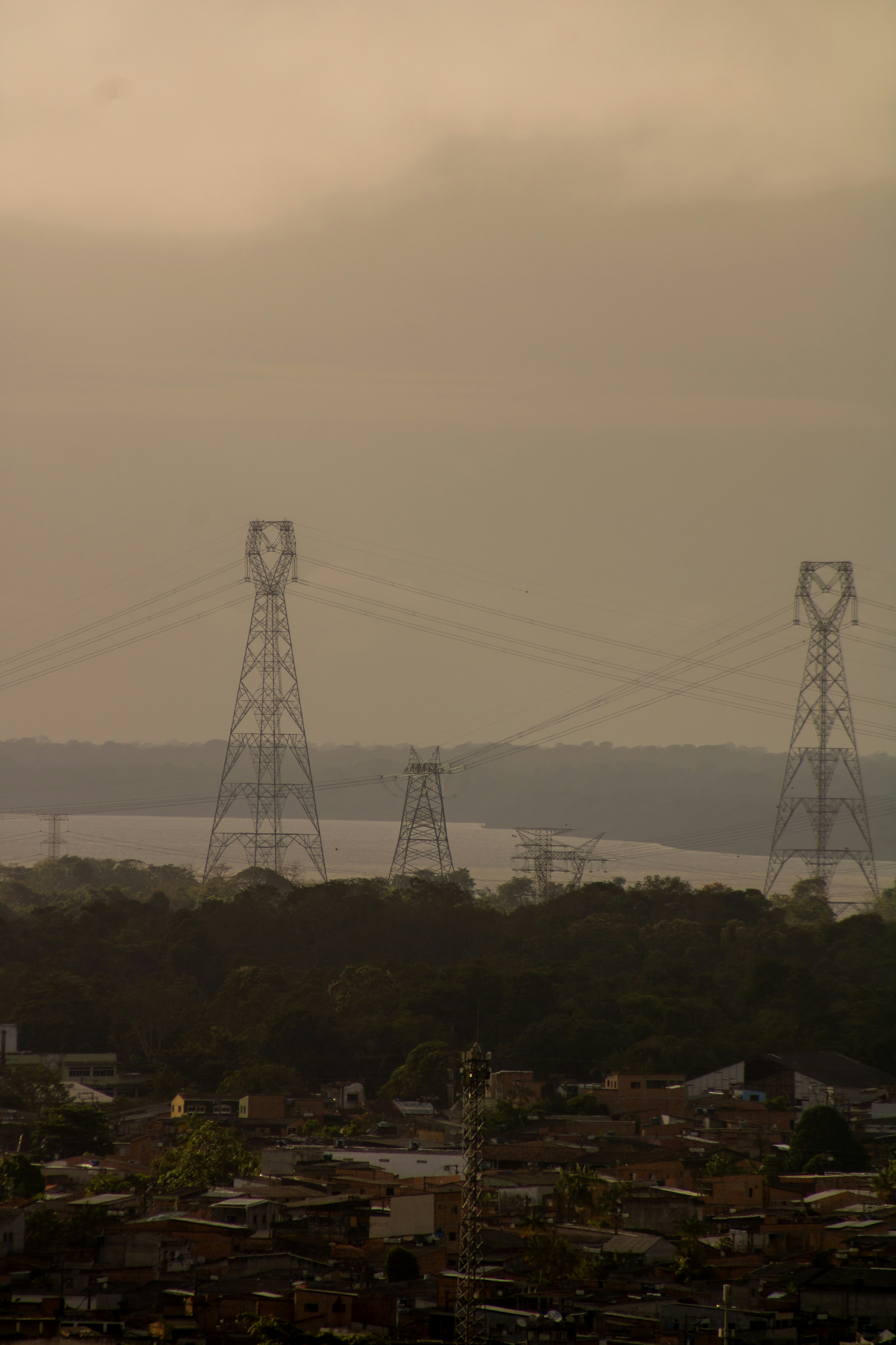 Wide shot of transmission towers and power lines above a suburban area at dusk, with a hazy sky and distant treeline.