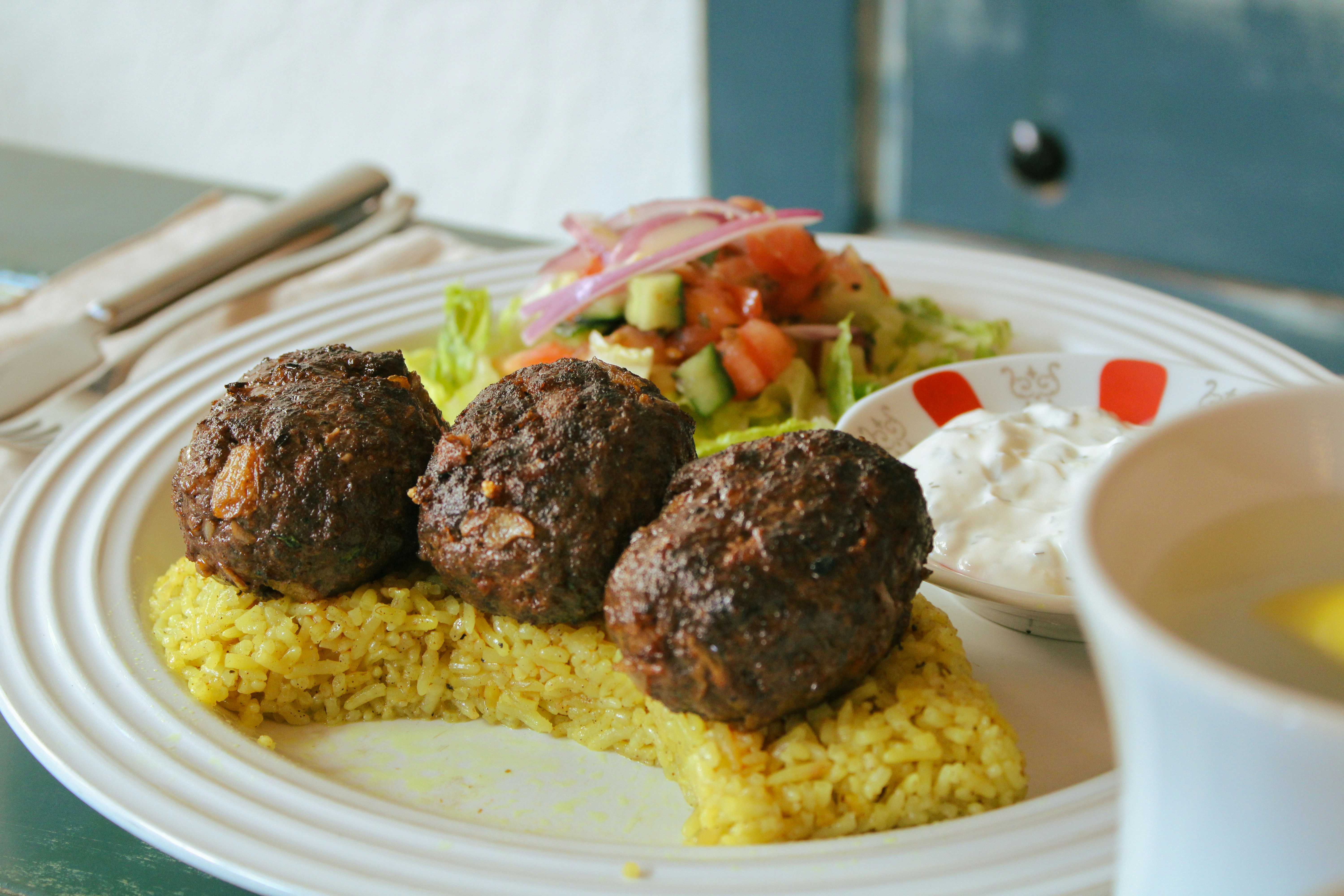 Serrano meatballs rest atop yellow saffron rice, accompanied by a small salad and a dollop of white sauce on a white plate.