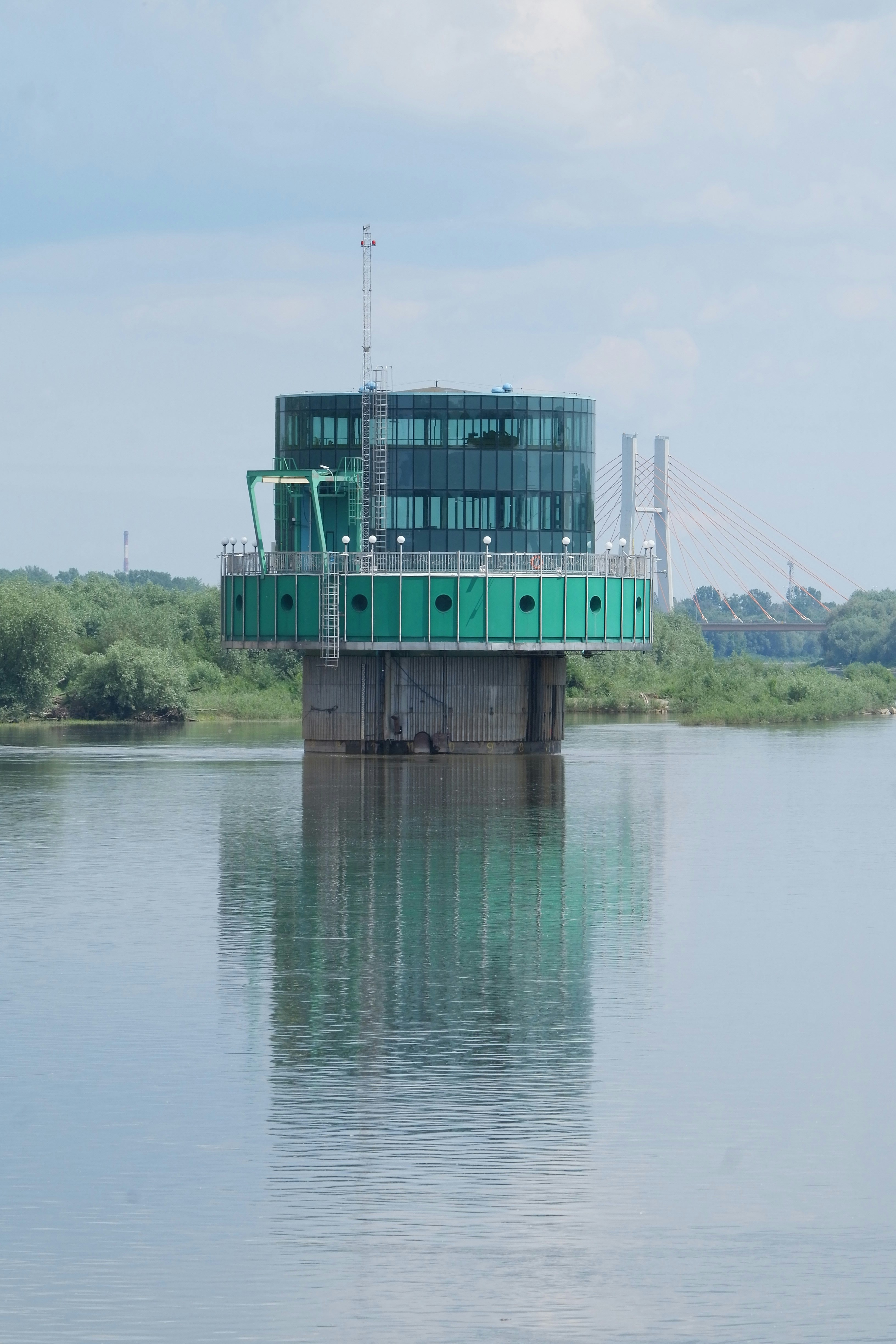 A green, multi-level structure rises from the water on a calm river, framed by distant trees and a blue sky; the scene emphasizes the architectural detail and reflections on the surface.