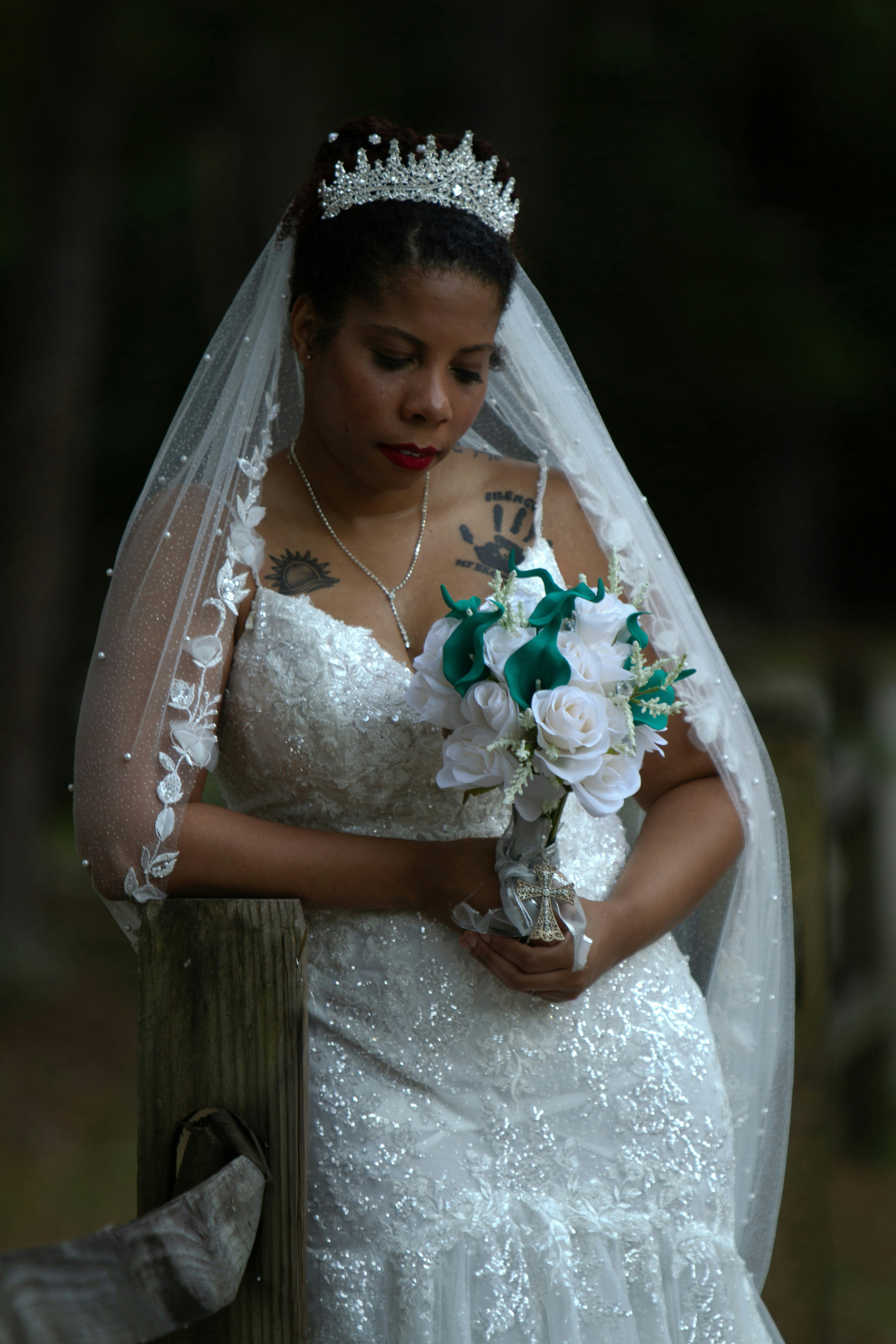 Bride in a jeweled tiara and sparkling wedding gown stands with a bouquet, veil draped over shoulders, hands clasped, outdoors. A portrait-like wedding moment.