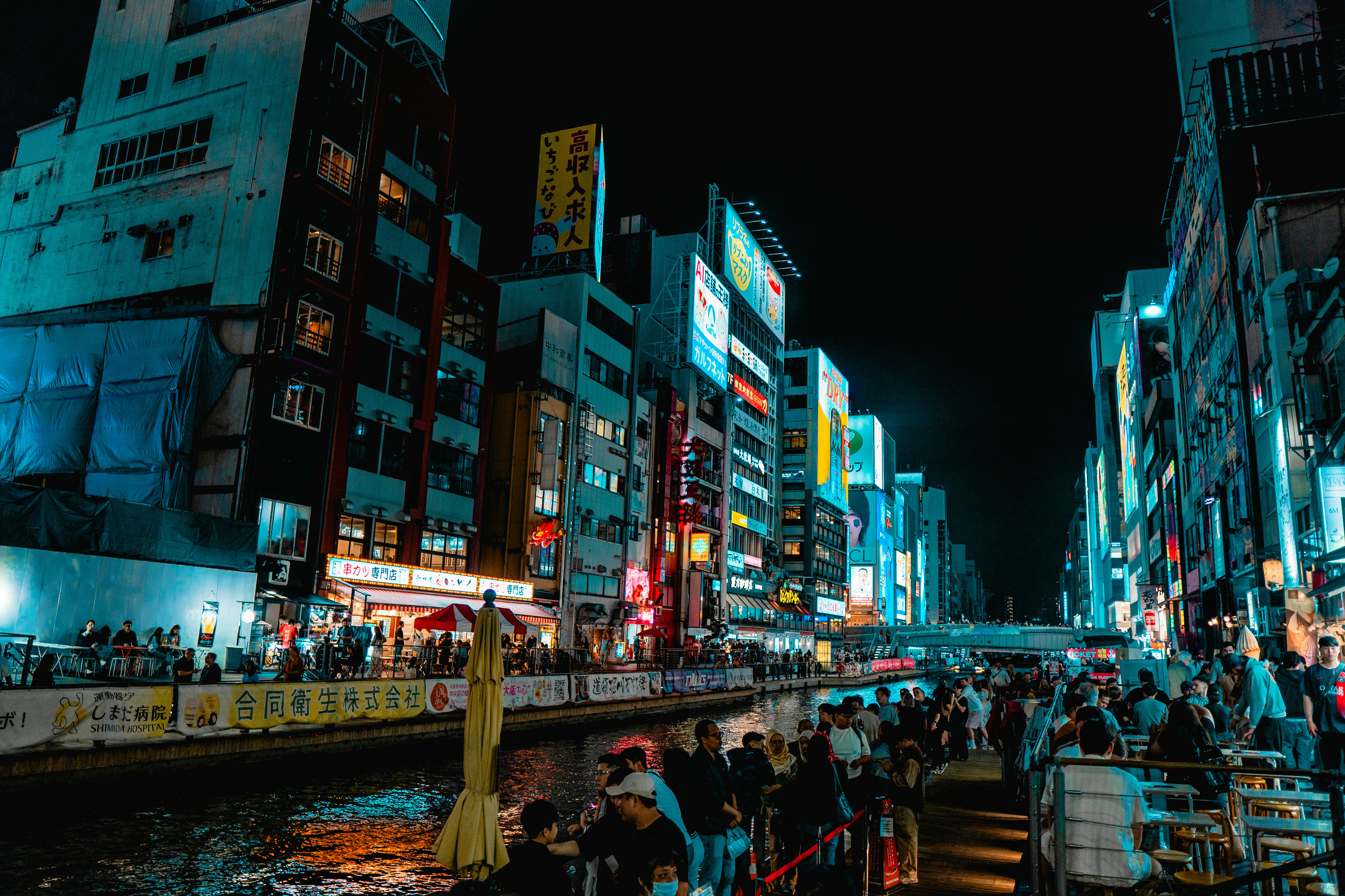 Nighttime canal cityscape with a crowded riverside promenade and illuminated signs along tall buildings; foreground platforms and boat silhouettes frame a lively urban scene.