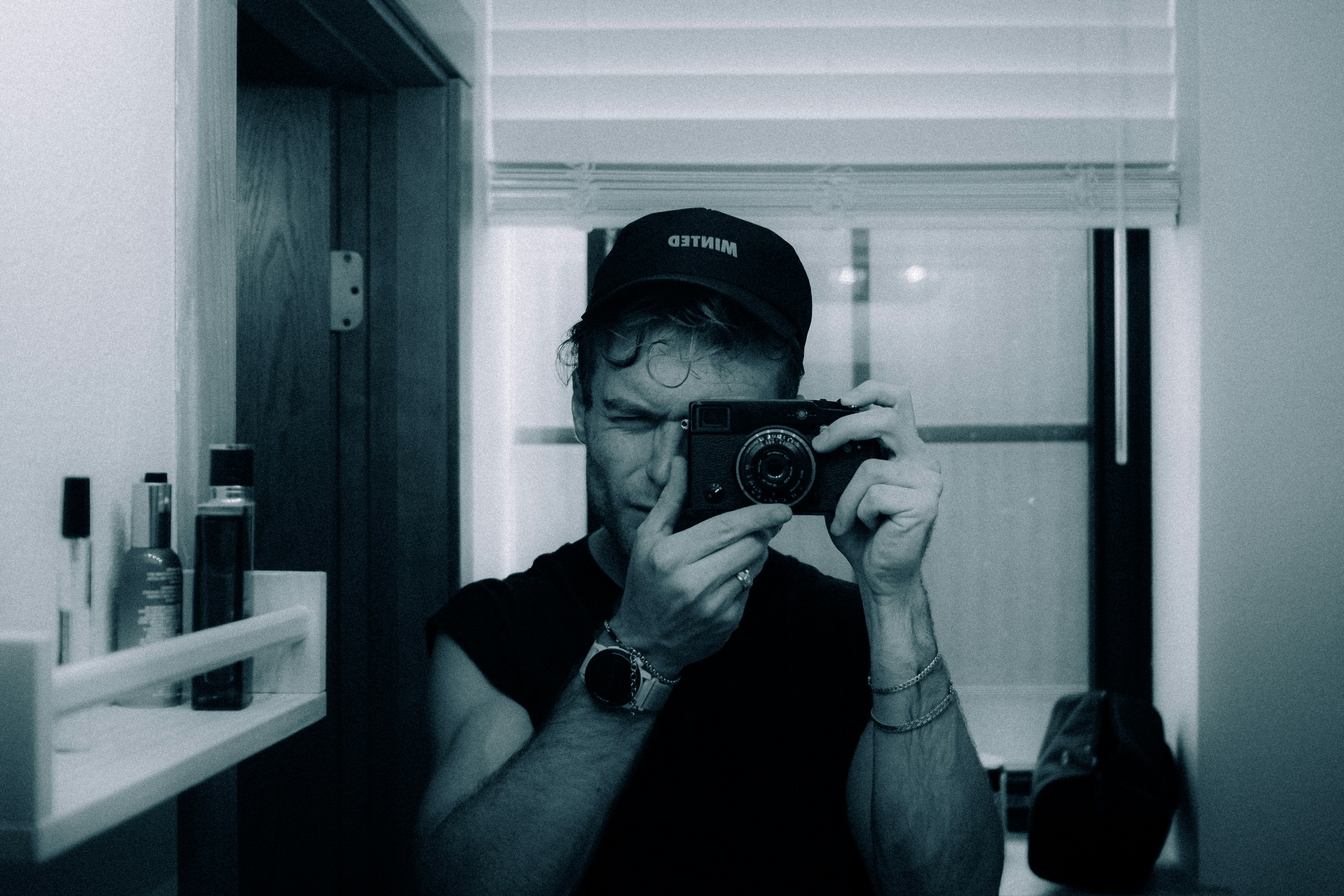 Portrait of a man taking a mirror selfie with a compact camera; window blinds and bathroom items visible in the background, rendered in a cool teal tone. The shot is a candid, with the subject centered and focused on the camera display.