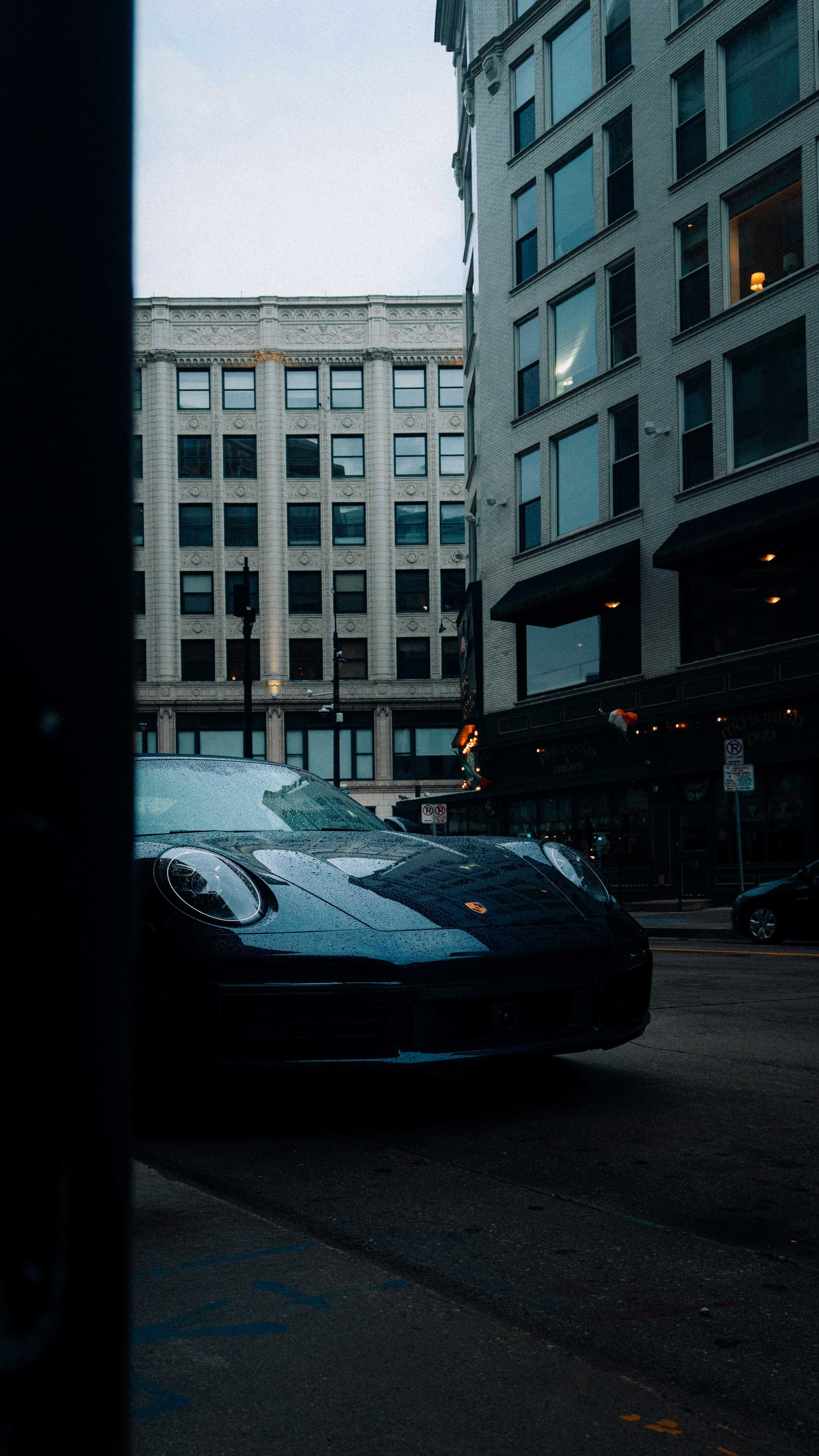 A blue Porsche sits at curb with tall city buildings framing the scene; the image emphasizes urban depth and moody lighting. It appears as a street-level capture, with the car as the focal point.