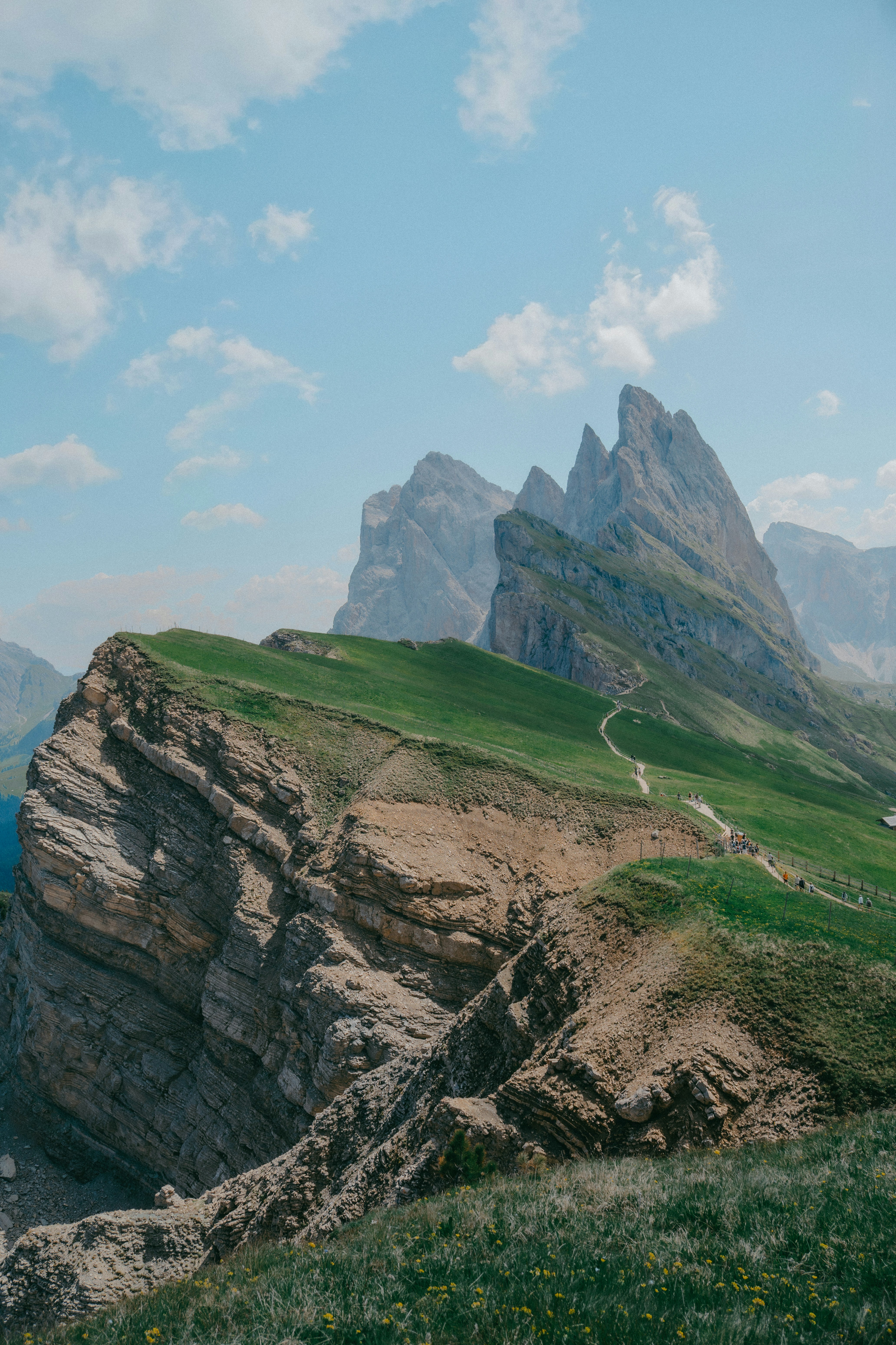 Sunlit coastal ridge winds toward jagged mountains as a narrow trail threads through grassy turf and rocky outcrops.