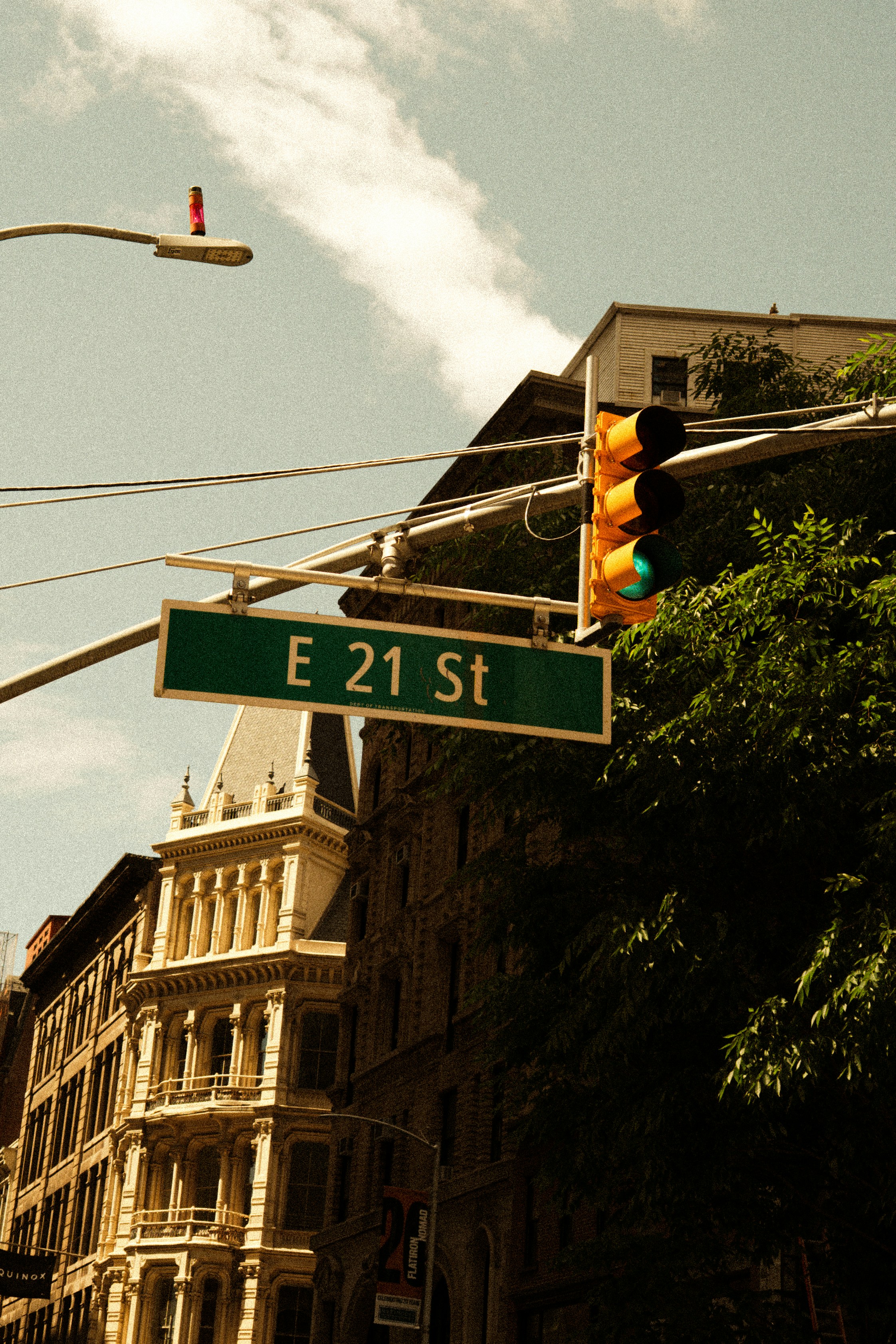 A city street scene featuring a green E 21 St sign suspended over a busy intersection against classic brick buildings and a traffic light; the composition highlights urban architecture and street infrastructure.