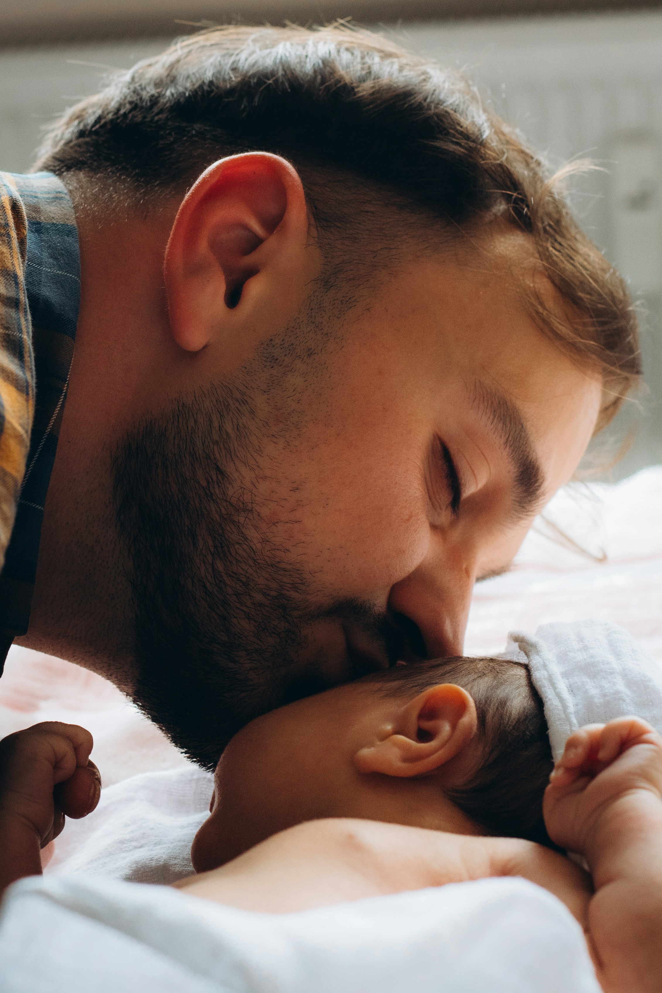 Close-up of a father kissing a newborn on the forehead, soft natural light highlighting intimate moment and skin tones. A candid, warm portrayal suitable for family photography; image depicts a real-life moment, not a staged illustration, with no logos visible.