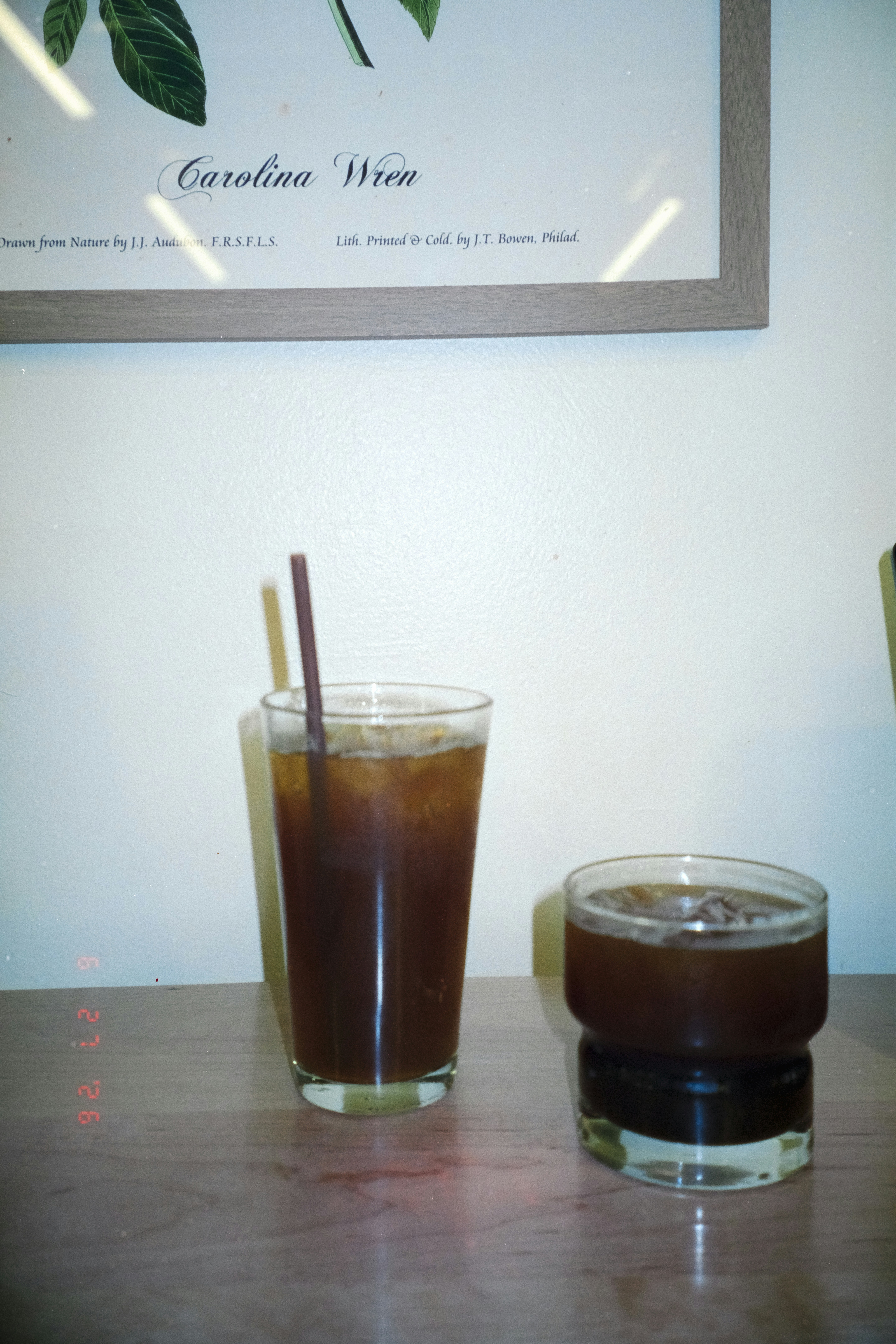 Two Glasses on a Wooden Table under Framed Botanical Art