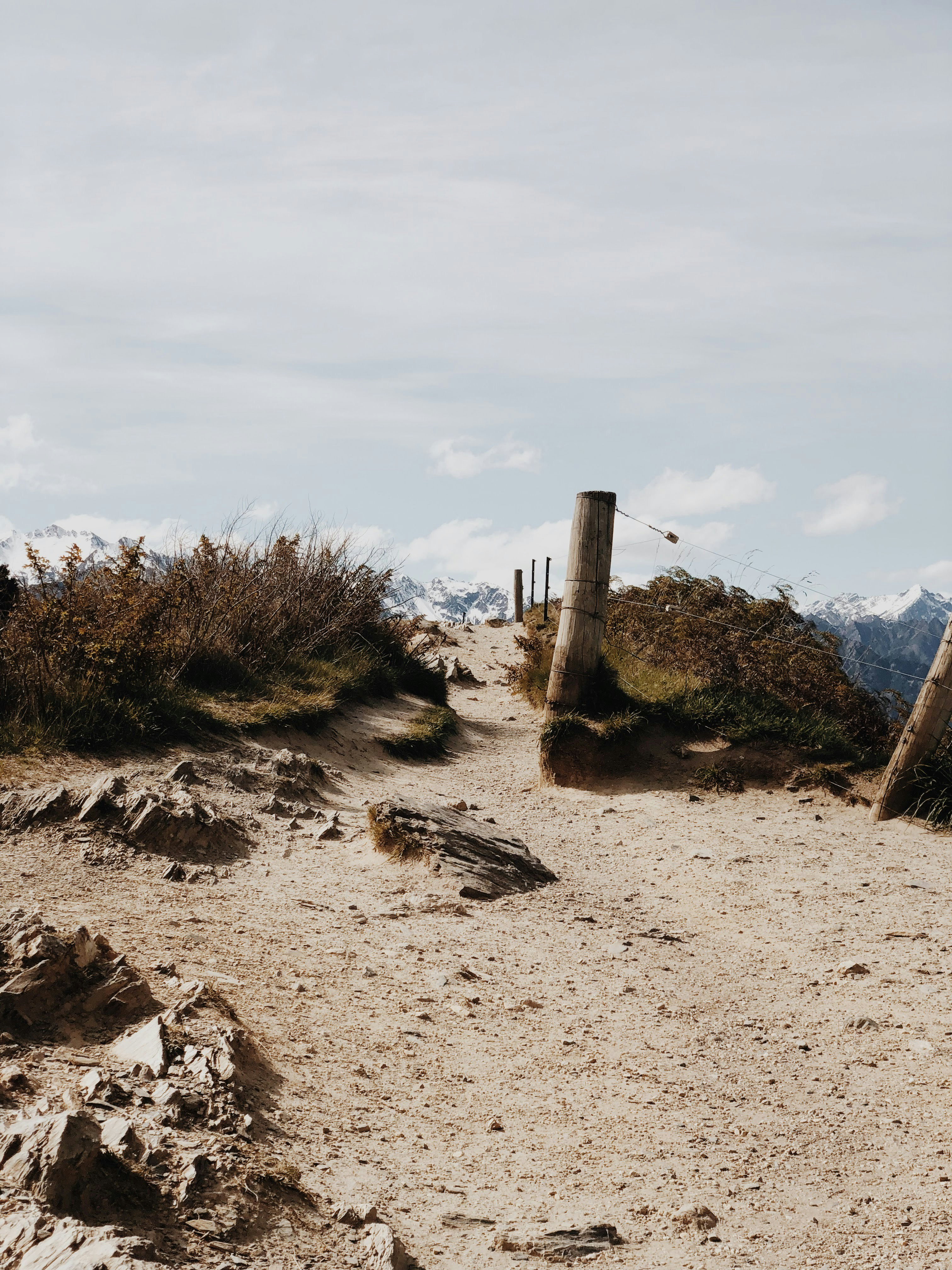 Dusty path along a ridge with weathered fenceposts leads toward distant snow-capped mountains; dry brush lines the trail, under a pale sky.