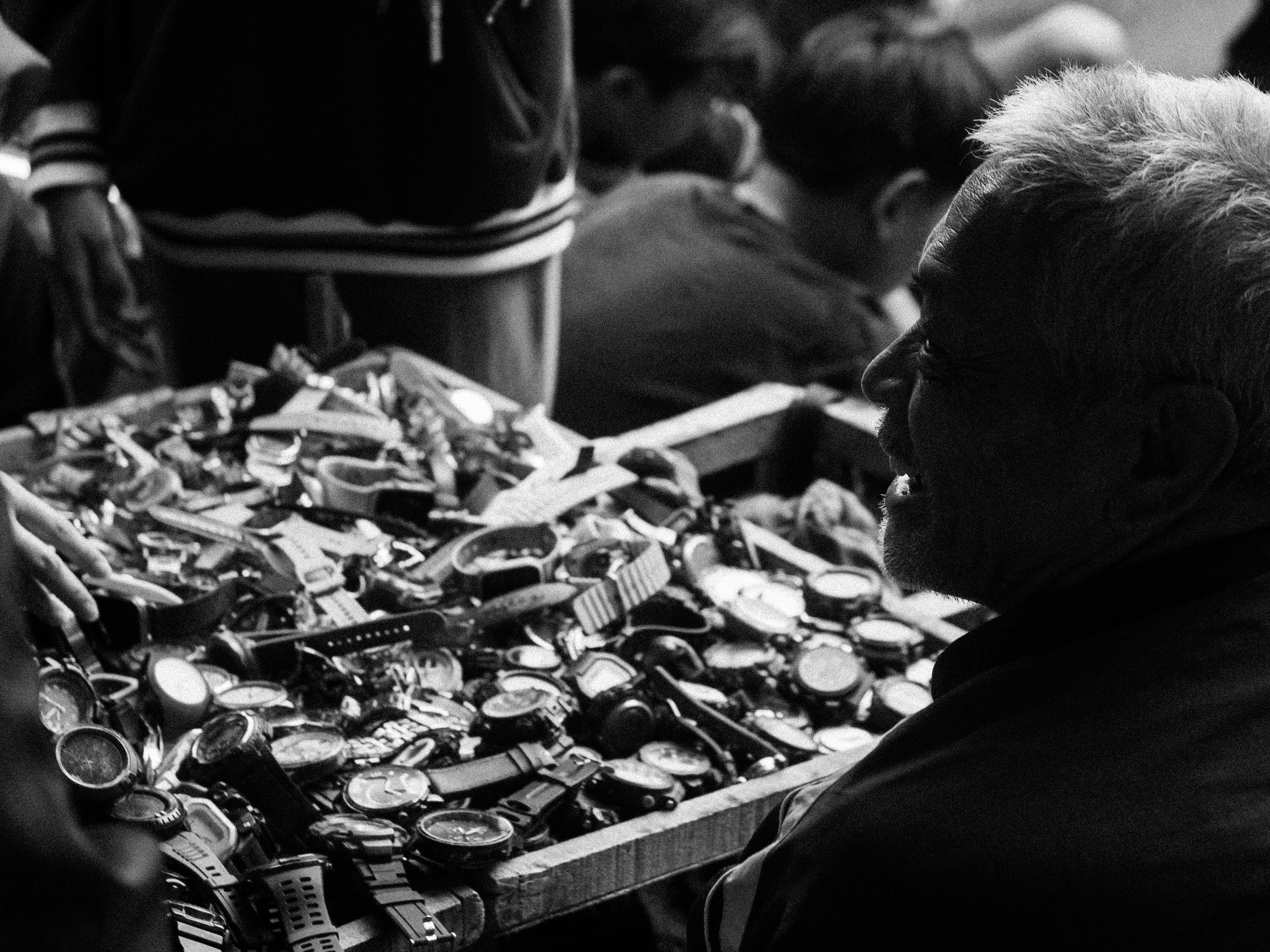 Close-up street scene of an elderly vendor among a table crowded with watches, lantern-like faces glinting in high-contrast light.