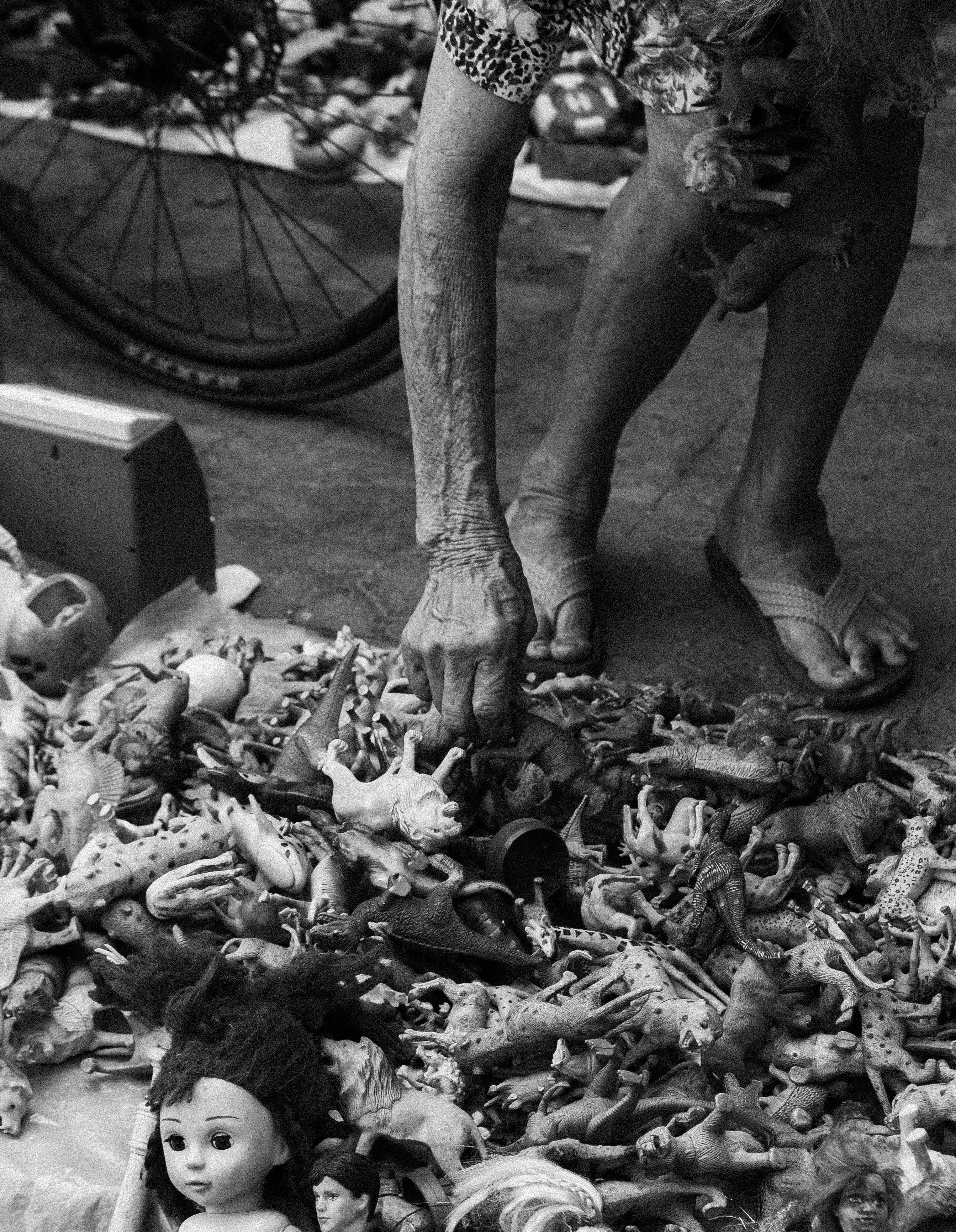 Row of small figurines spilled on the ground with a pair of hands and bare feet selecting or arranging them; a bicycle wheel and toolbox appear in the background, suggesting a street market scene with discarded toys.