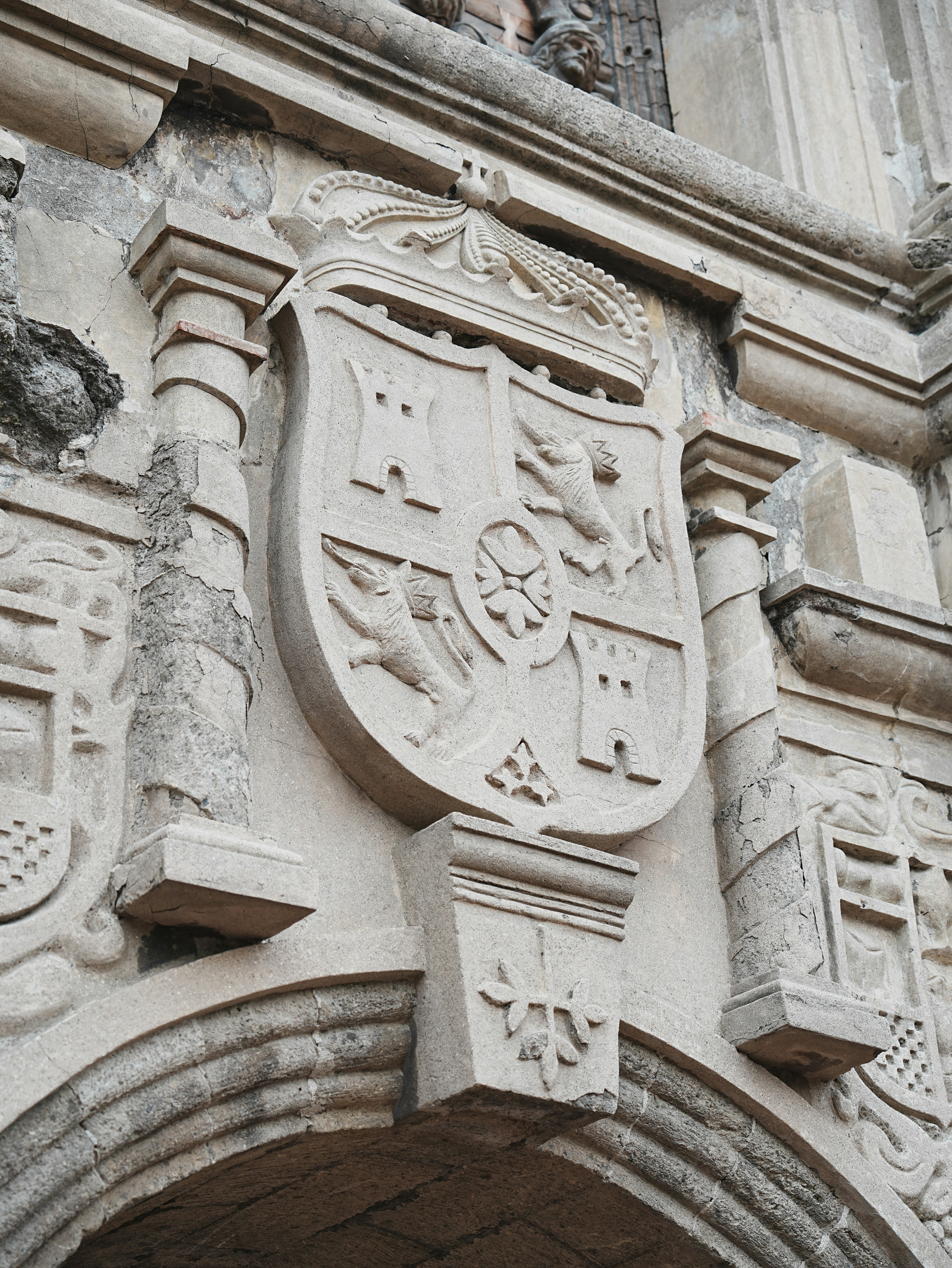 Intricately carved shield crest set into a weathered stone arch, featuring central floral motif and heraldic elements on an ornate architectural facade.