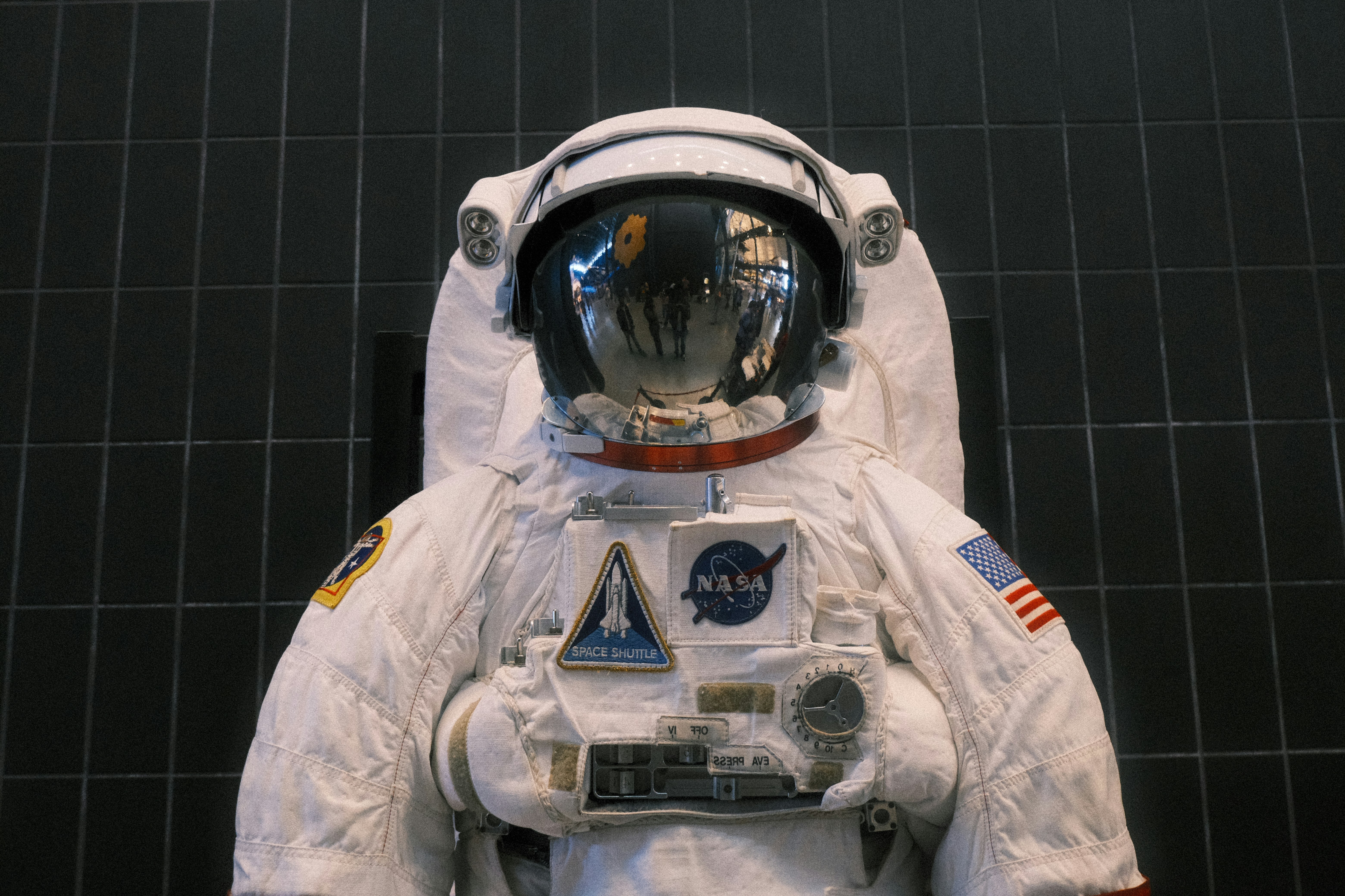 White spacesuit with NASA patches and reflective helmet visor stands against a dark panel, capturing a ceremonial, still pose. The image is likely a display or exhibit photograph.