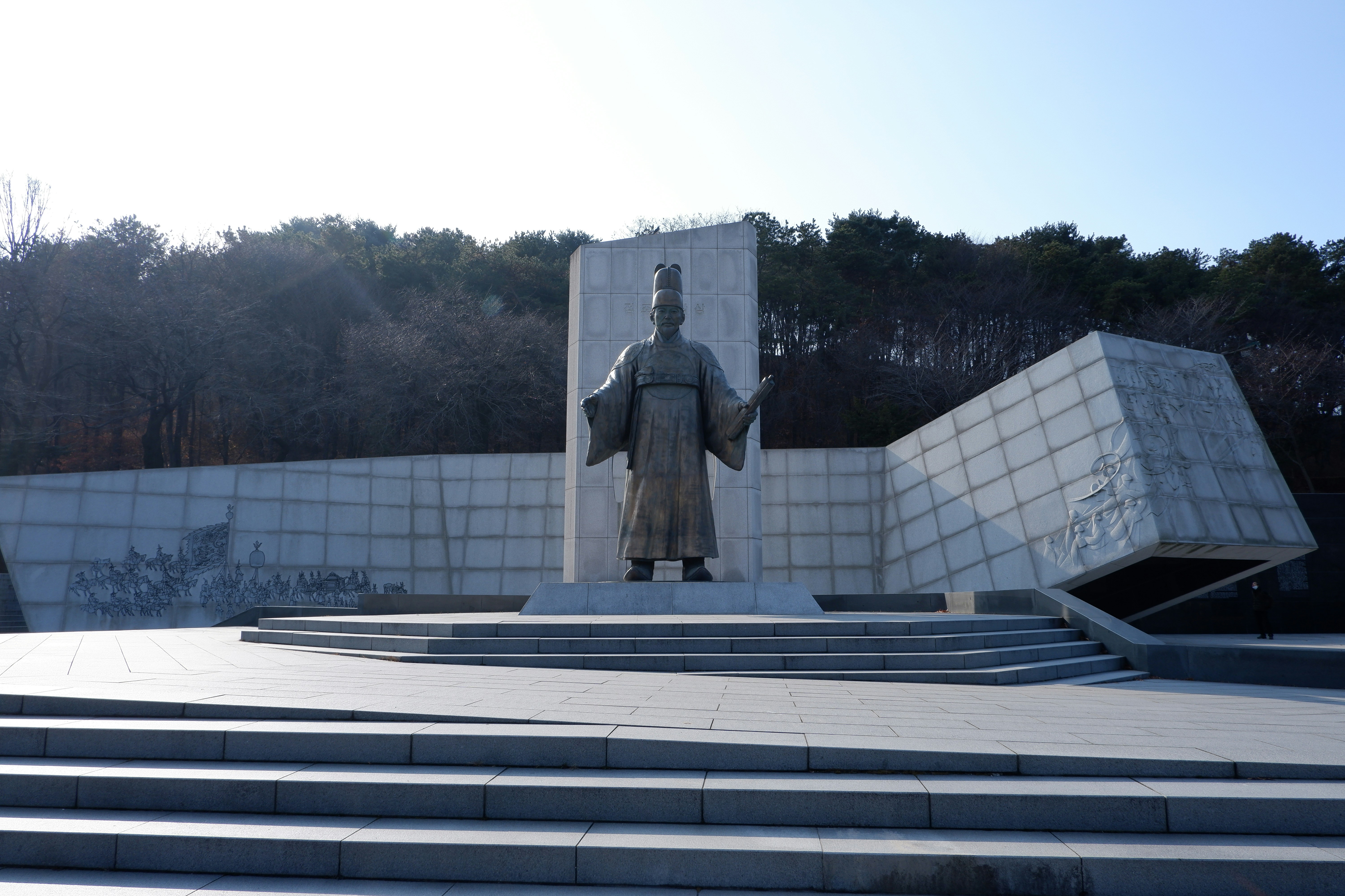 Bronze Statue of King Jeongjo, located within the Hwaseong Fortress in Suwon, South Korea. King Jeongjo was a notable ruler of the Joseon Dynasty known for building the fortress to protect his father's tomb and establish a new city. The surrounding area features stone walls engraved with historical information about the fortress and the king's achievements.