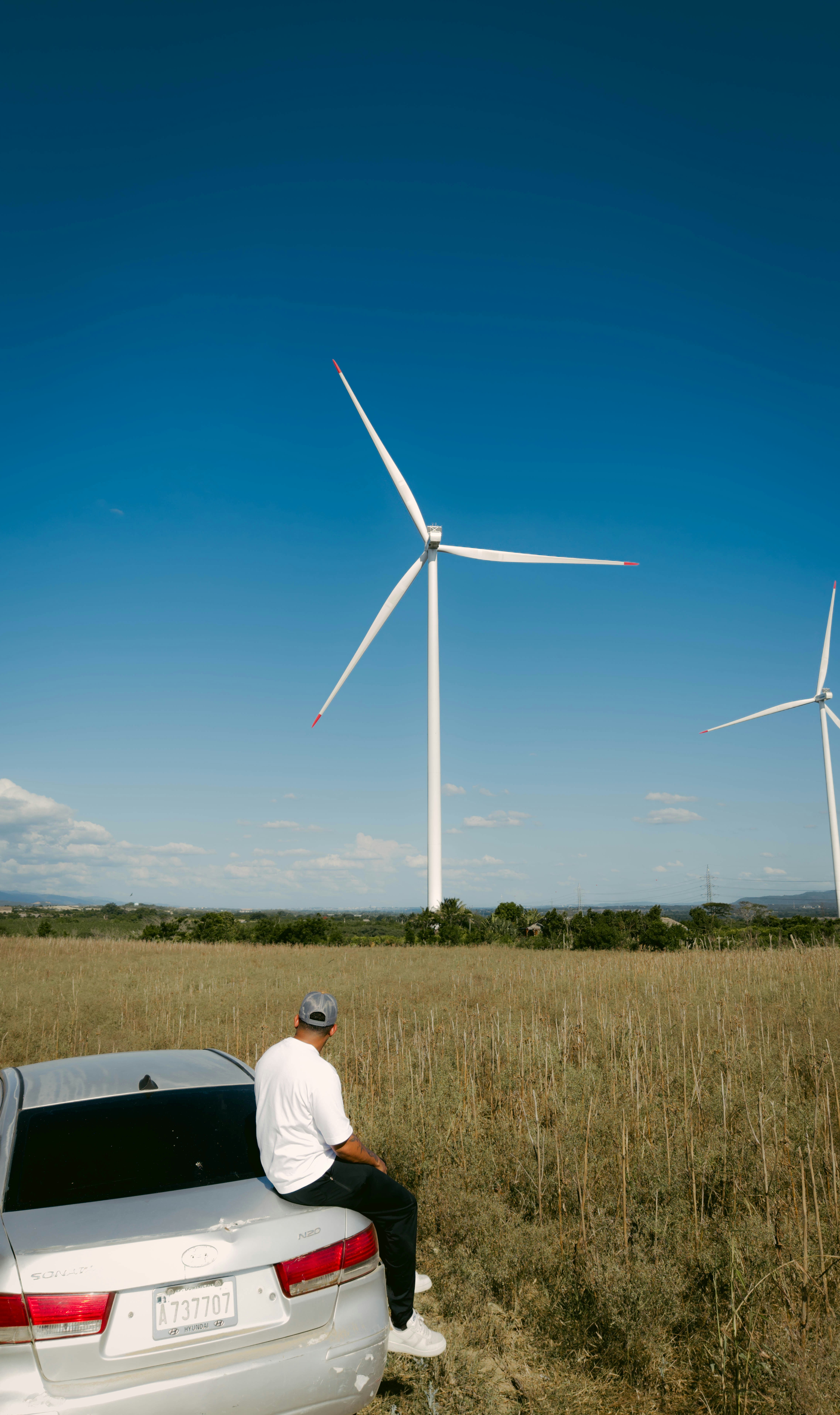Man sits on the trunk of a parked car in a golden field, with wind turbines towering in the background under a clear blue sky.