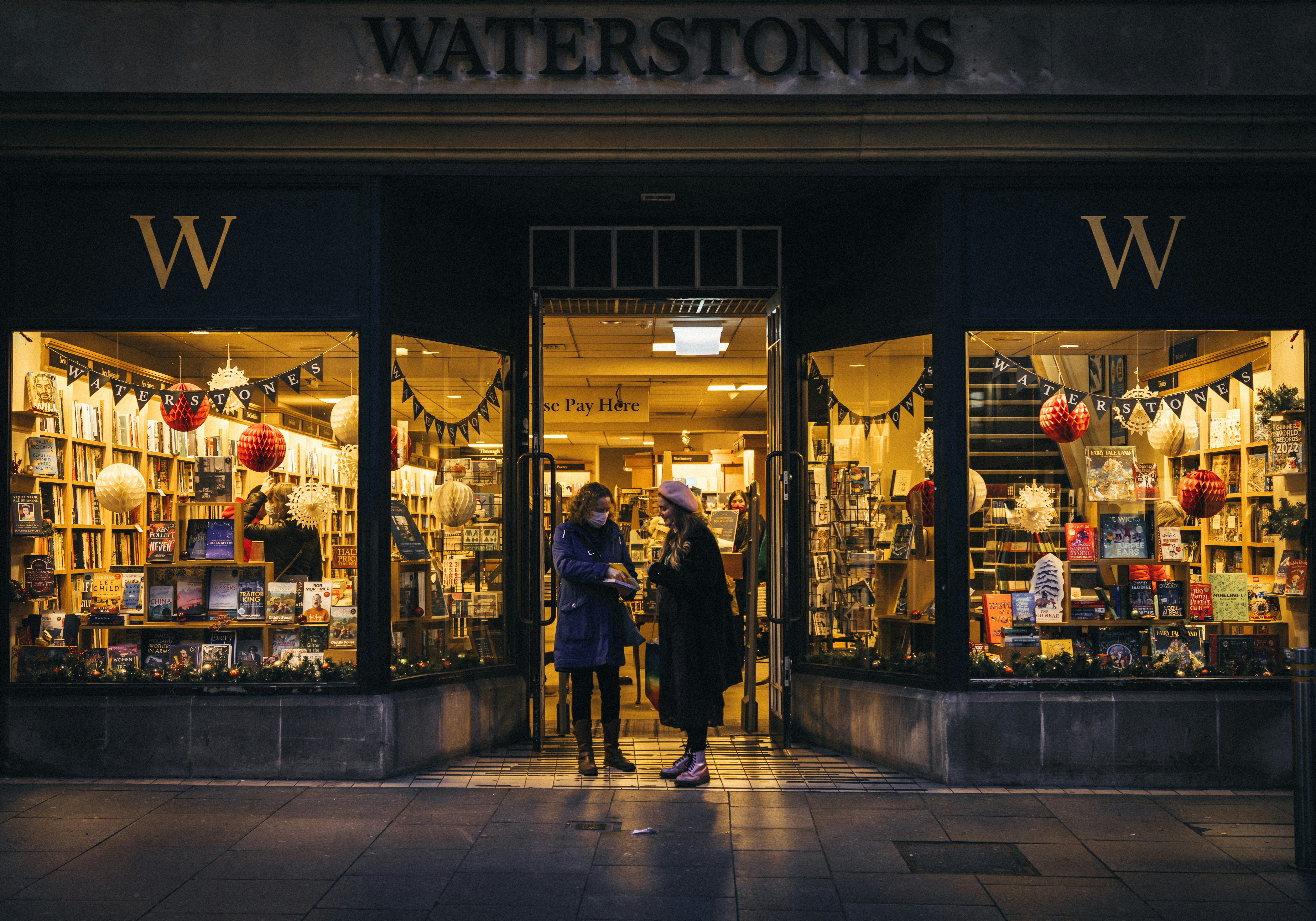 Two shoppers in a warmly lit bookstore window at dusk