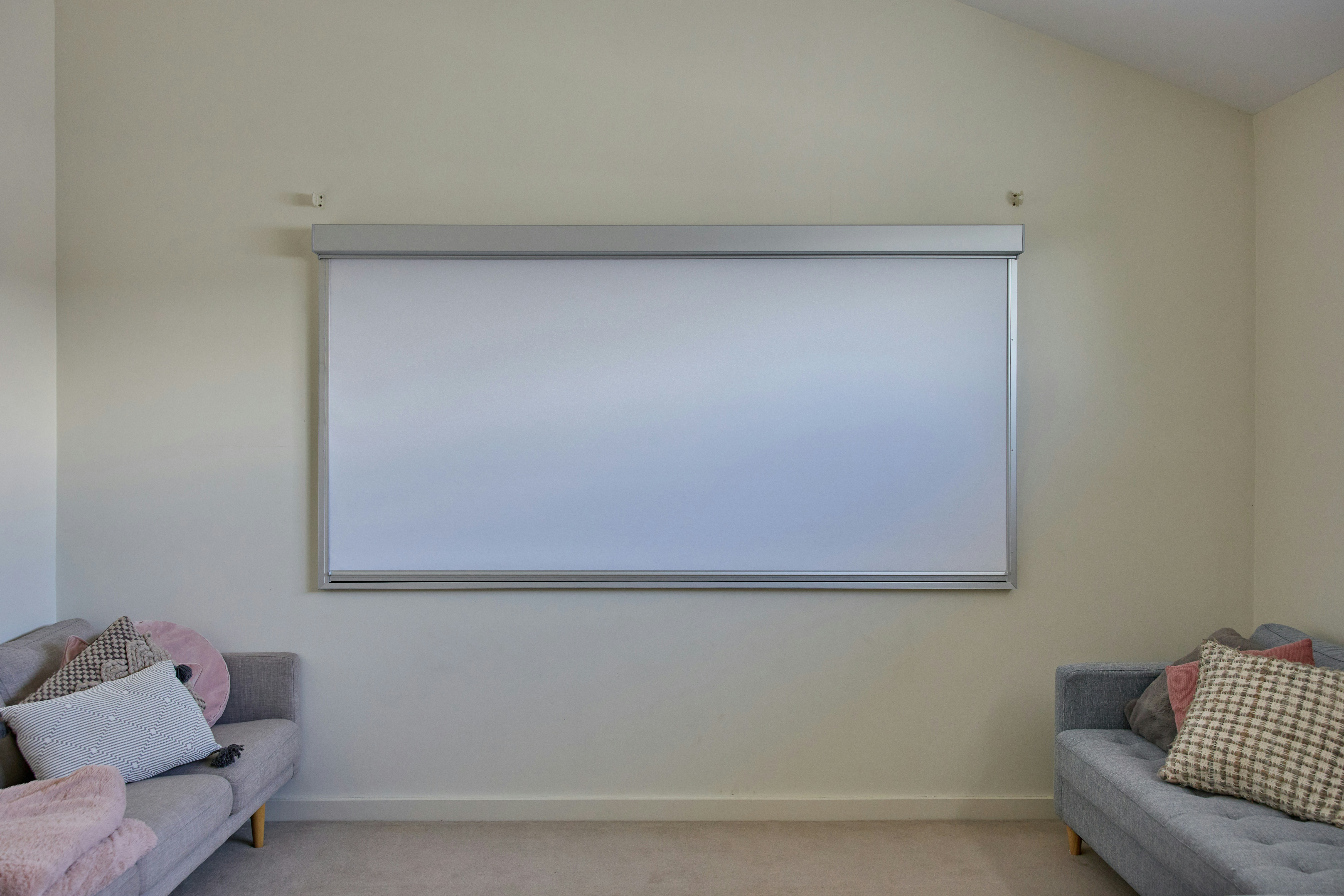 Soft-lit living room with a large, horizontally oriented window blind centered on a pale wall, flanked by two gray seating arrangements and patterned cushions.