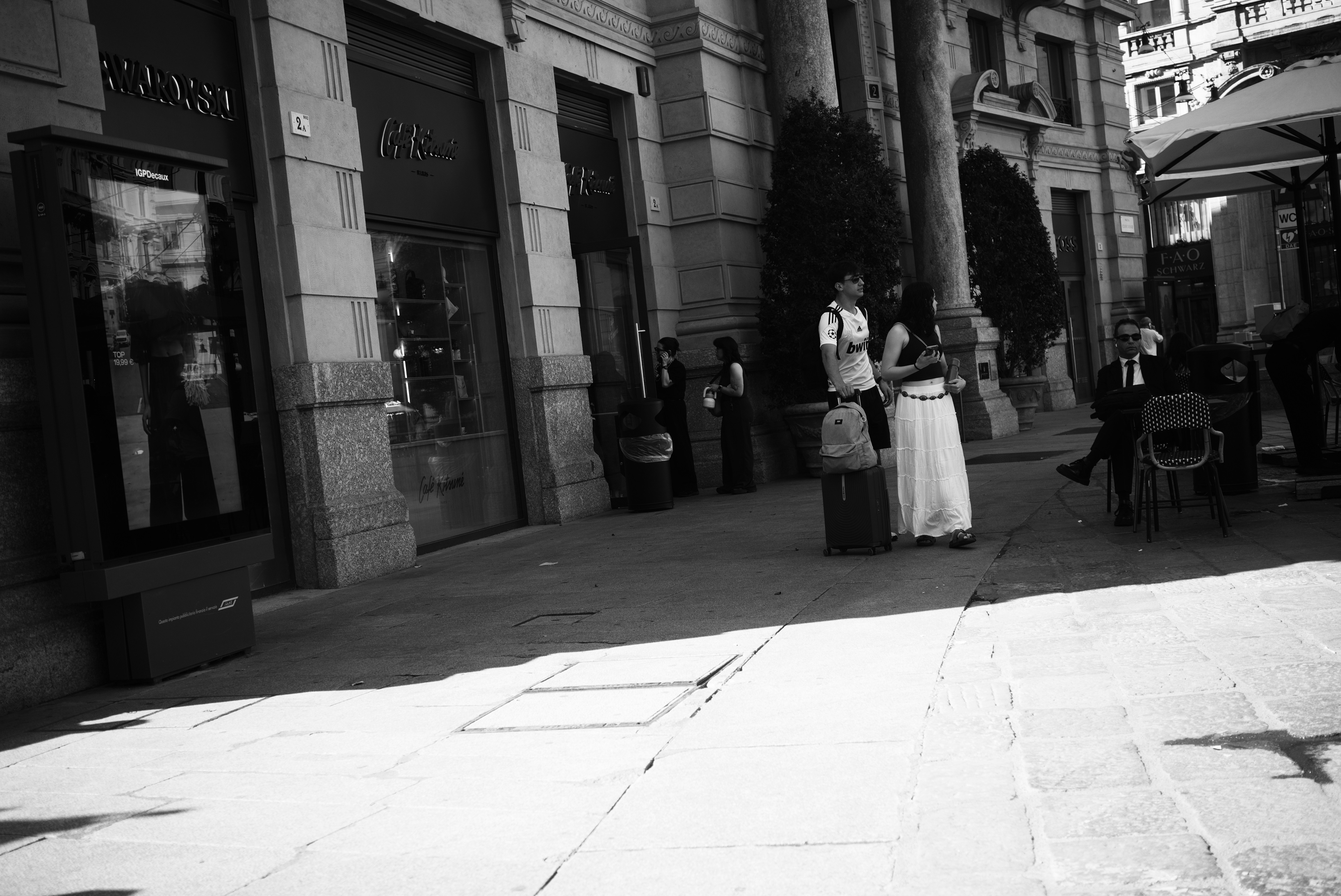 A grayscale urban scene shows a couple near a cafe under an umbrella, with passersby and storefronts lining a sunny sidewalk.