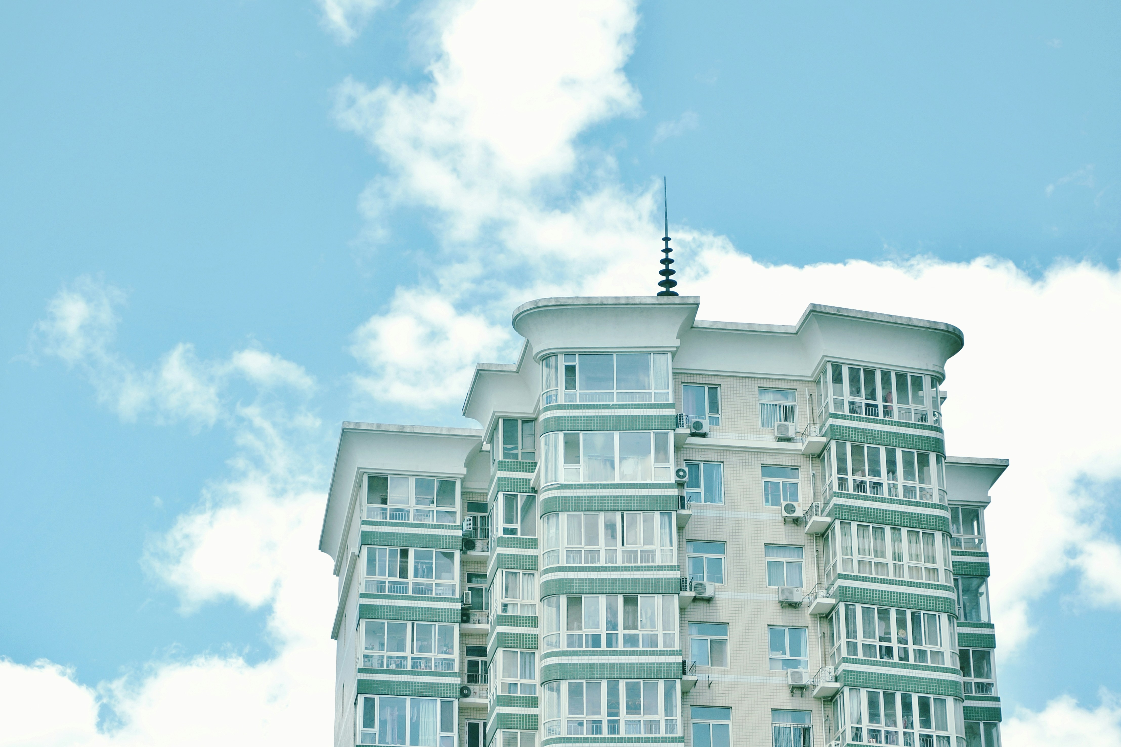 A pale, multi‑tiered residential tower rises into a bright blue sky with scattered clouds, the building's glass balconies catching soft light. The scene emphasizes urban verticality and modern architecture.