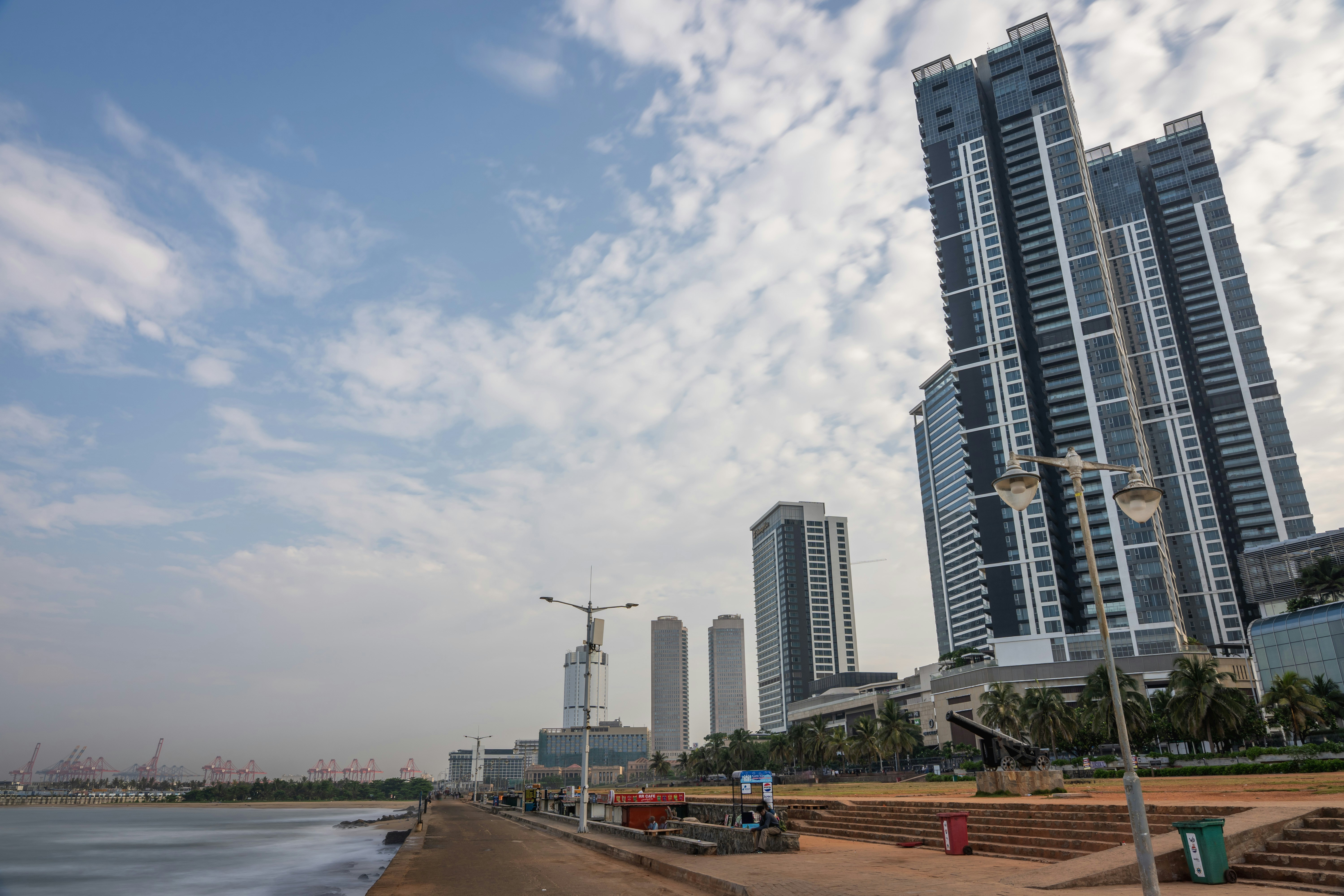 Low-angle view along a coastal promenade with modern high-rise buildings rising over a beach and calm water, capturing the urban shoreline and promenade activity.