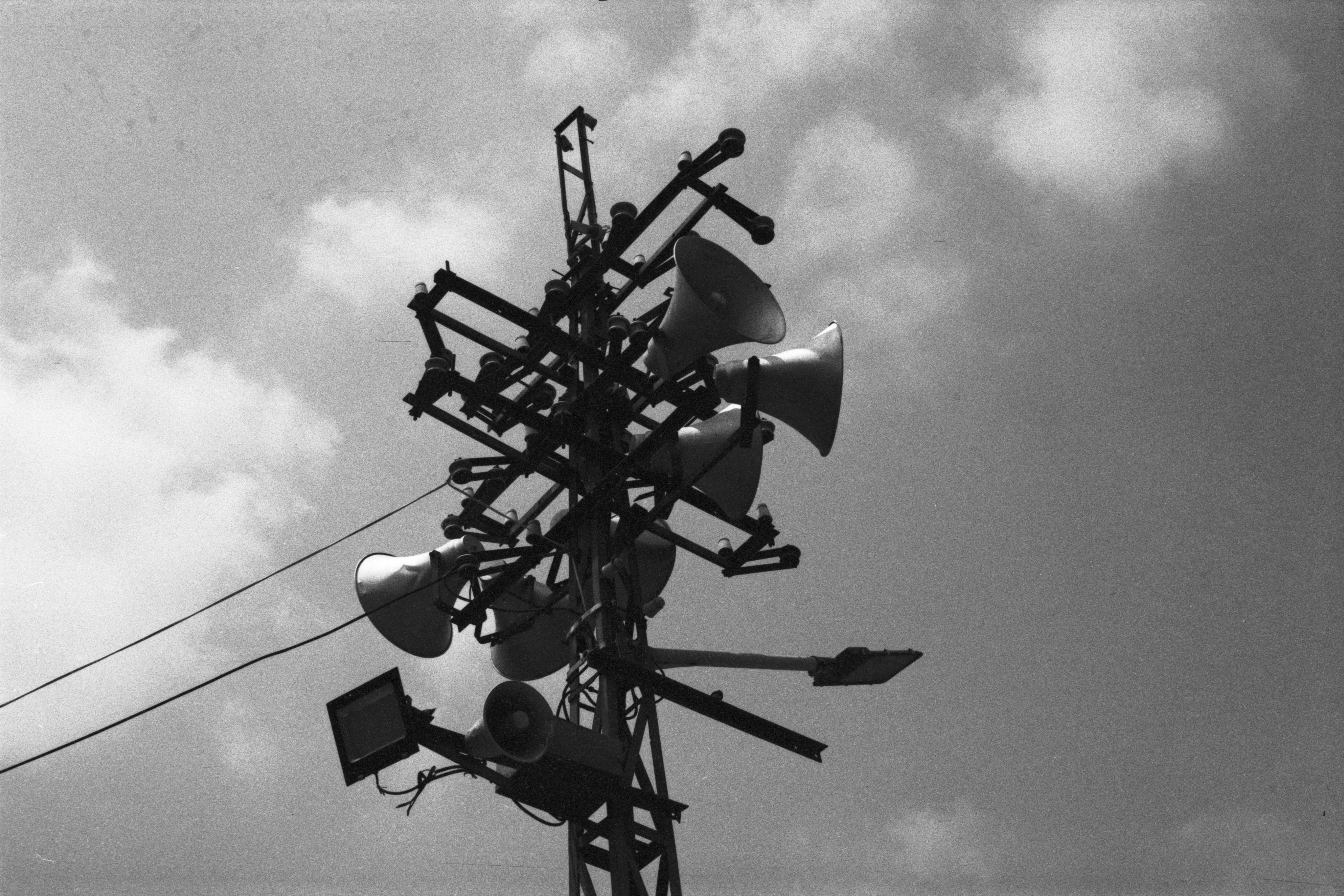 Industrial mast with multiple loudspeakers and fixtures rising toward a partly cloudy sky, silhouetted against light clouds.