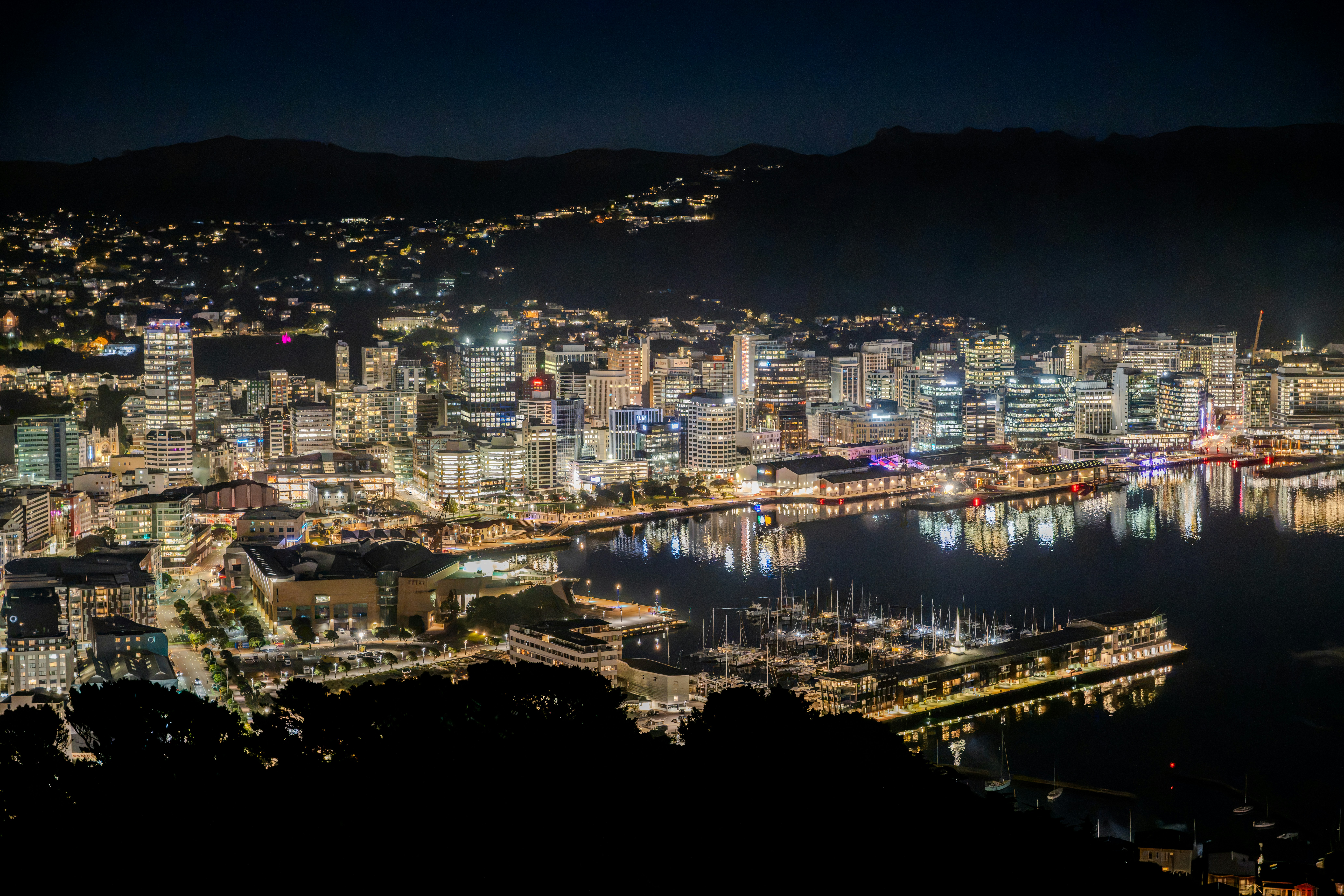 Nighttime cityscape along the harbor with illuminated skyline and reflections on the water.