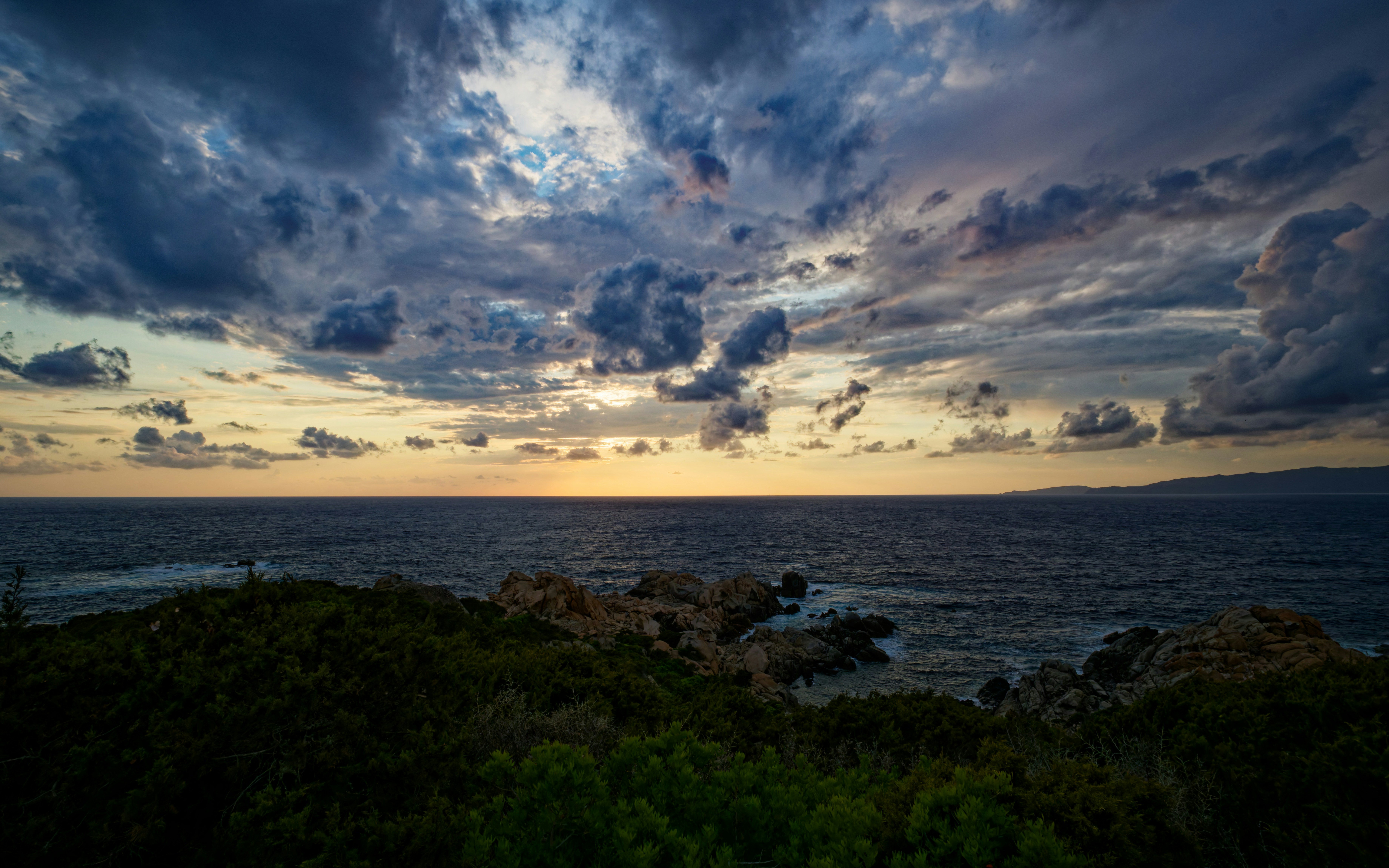 Coastal rocks and low-lying shrubbery frame a calm ocean at sunset, with dramatic clouds catching warm light. A landscape scene suitable for a serene nature display.