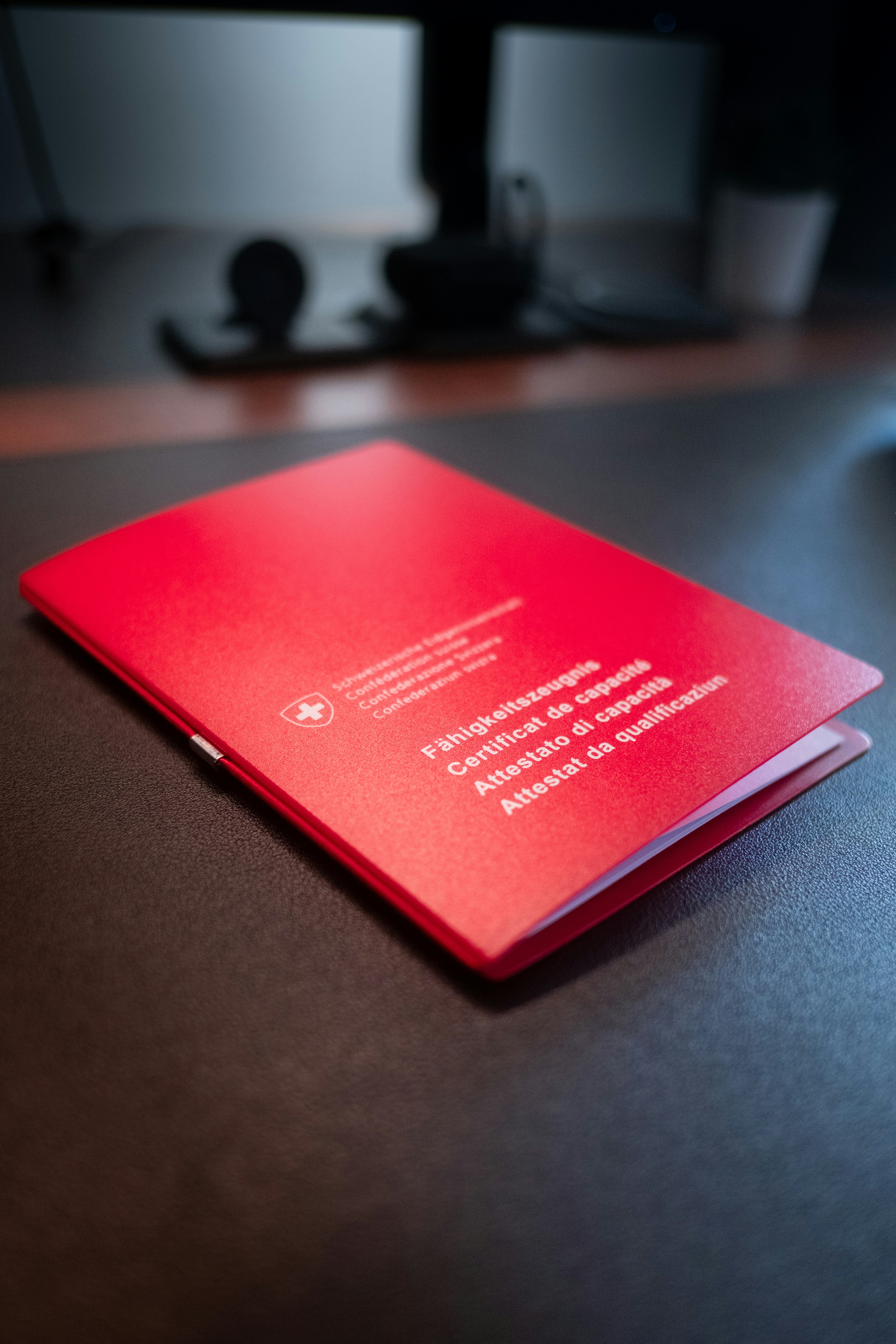 Red passport lies on a dark desk with blurred monitor and camera gear in the background, emphasizing texture and color. A logoed booklet under soft light.