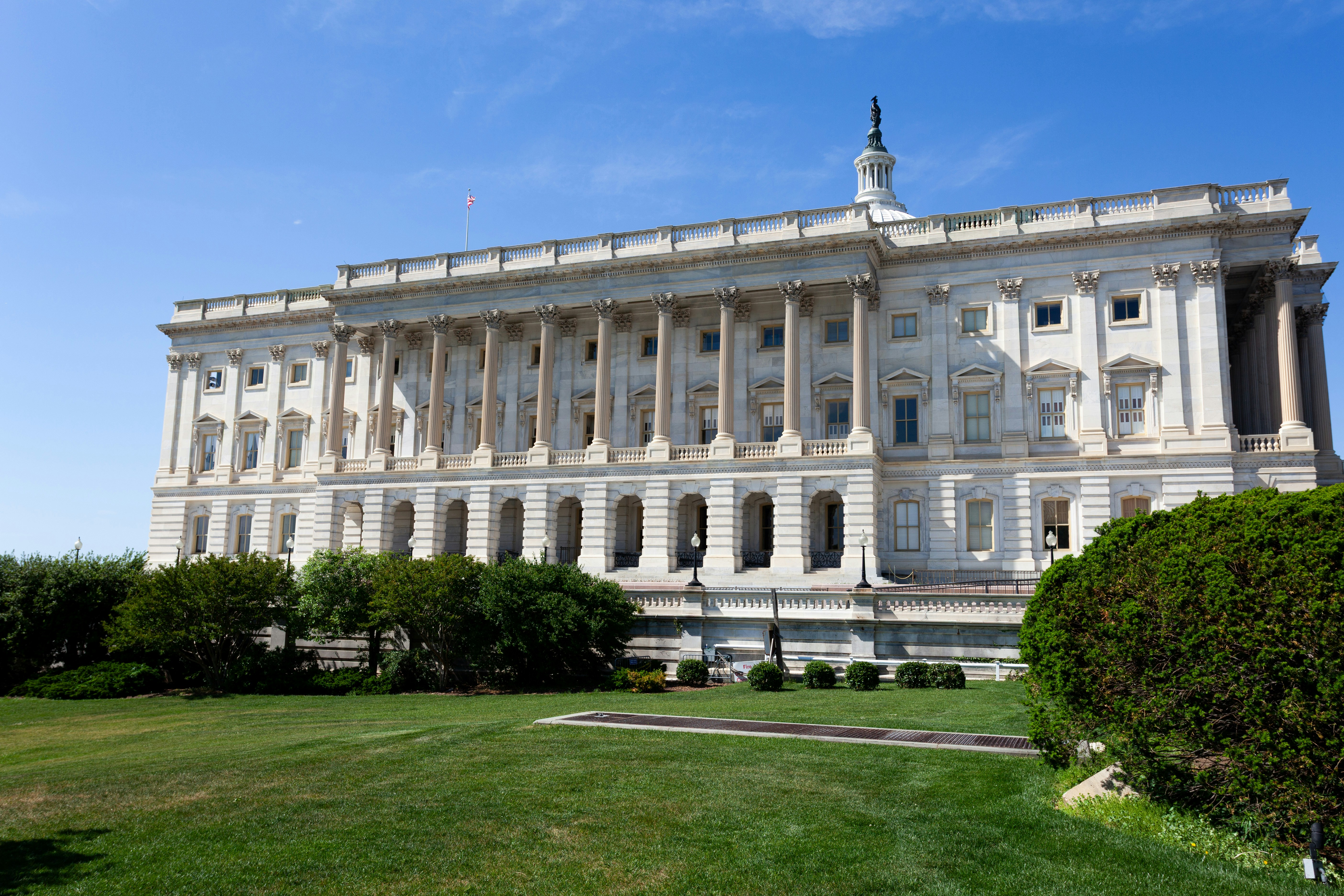 A clear, sunny day view of the exterior facade of the United States Capitol building in Washington, D.C., featuring its neoclassical architecture, columns, and surrounding green lawn.