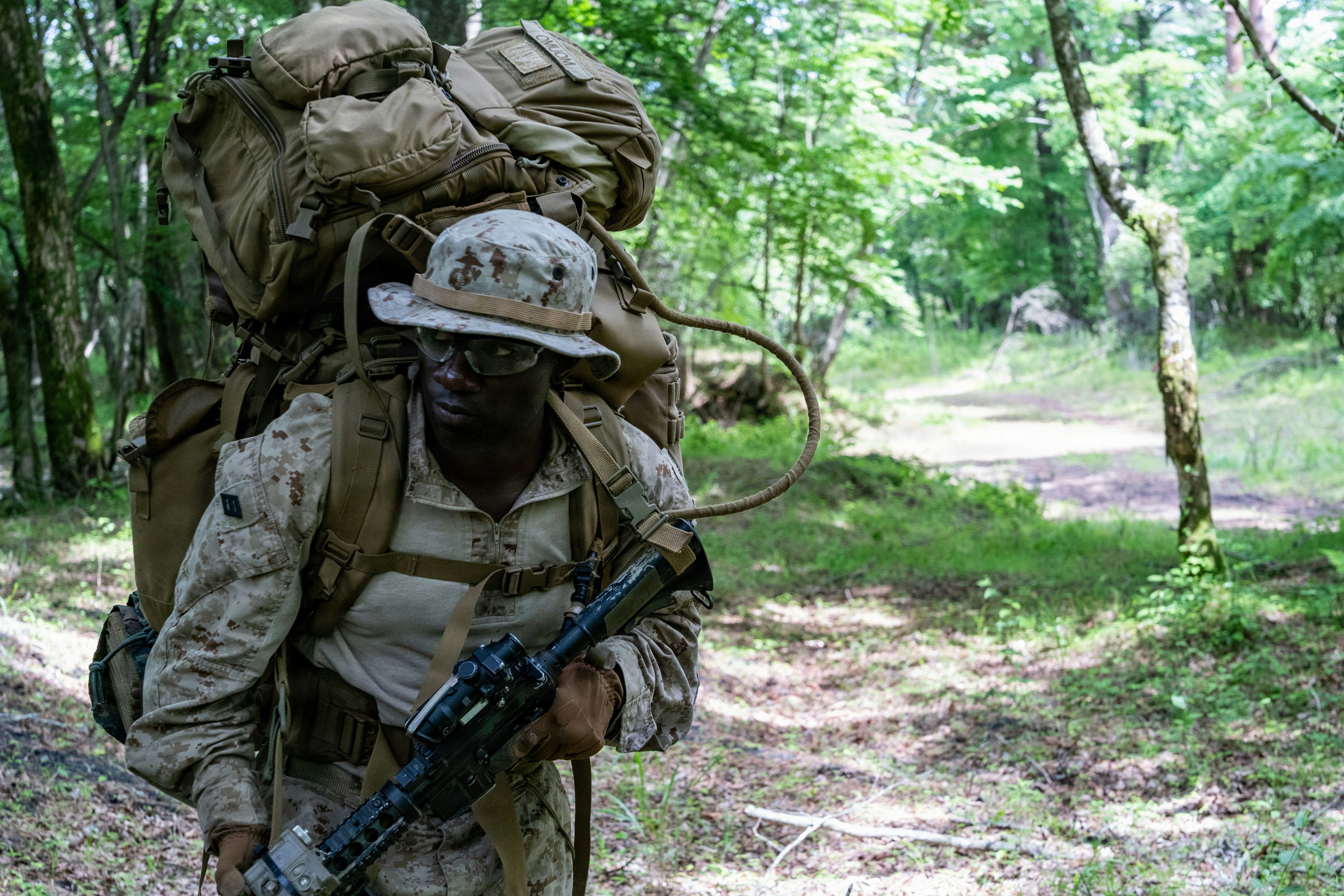 Camouflaged soldier in full gear moves through a woodland trail with a rifle slung, backpack secured, under dappled light.