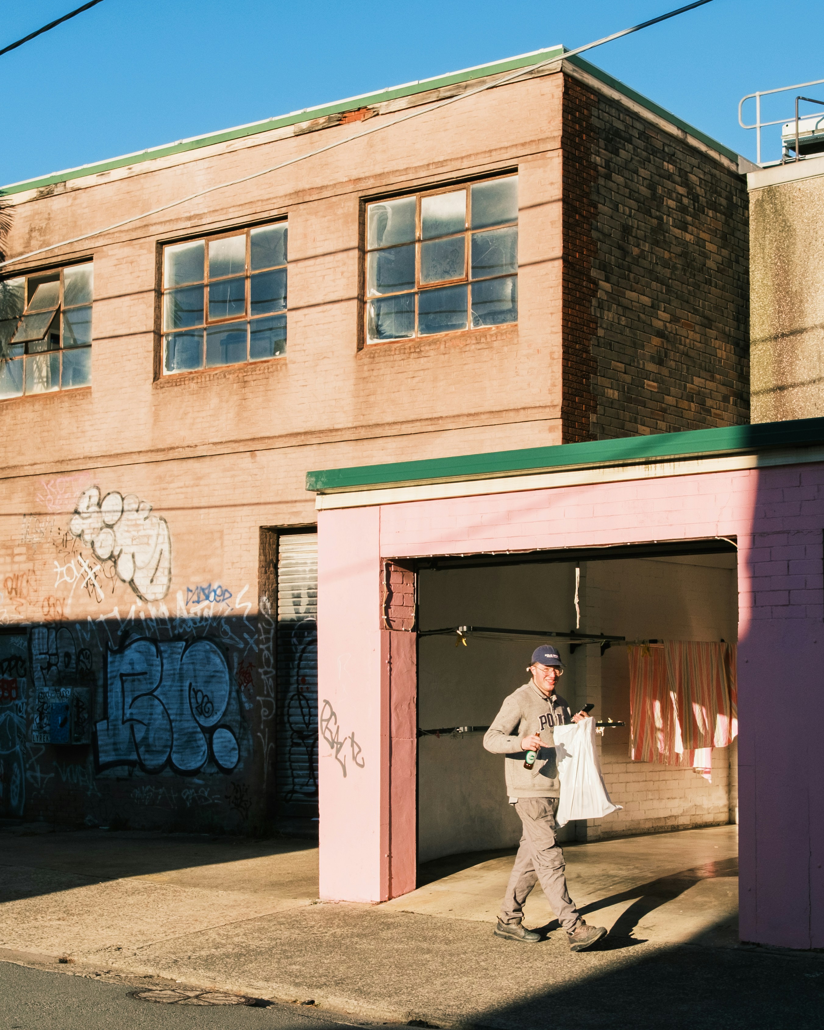 A man in neutral clothing walks under a pink-painted archway, laundry hanging nearby, with a graffiti-drenched brick wall in the background.