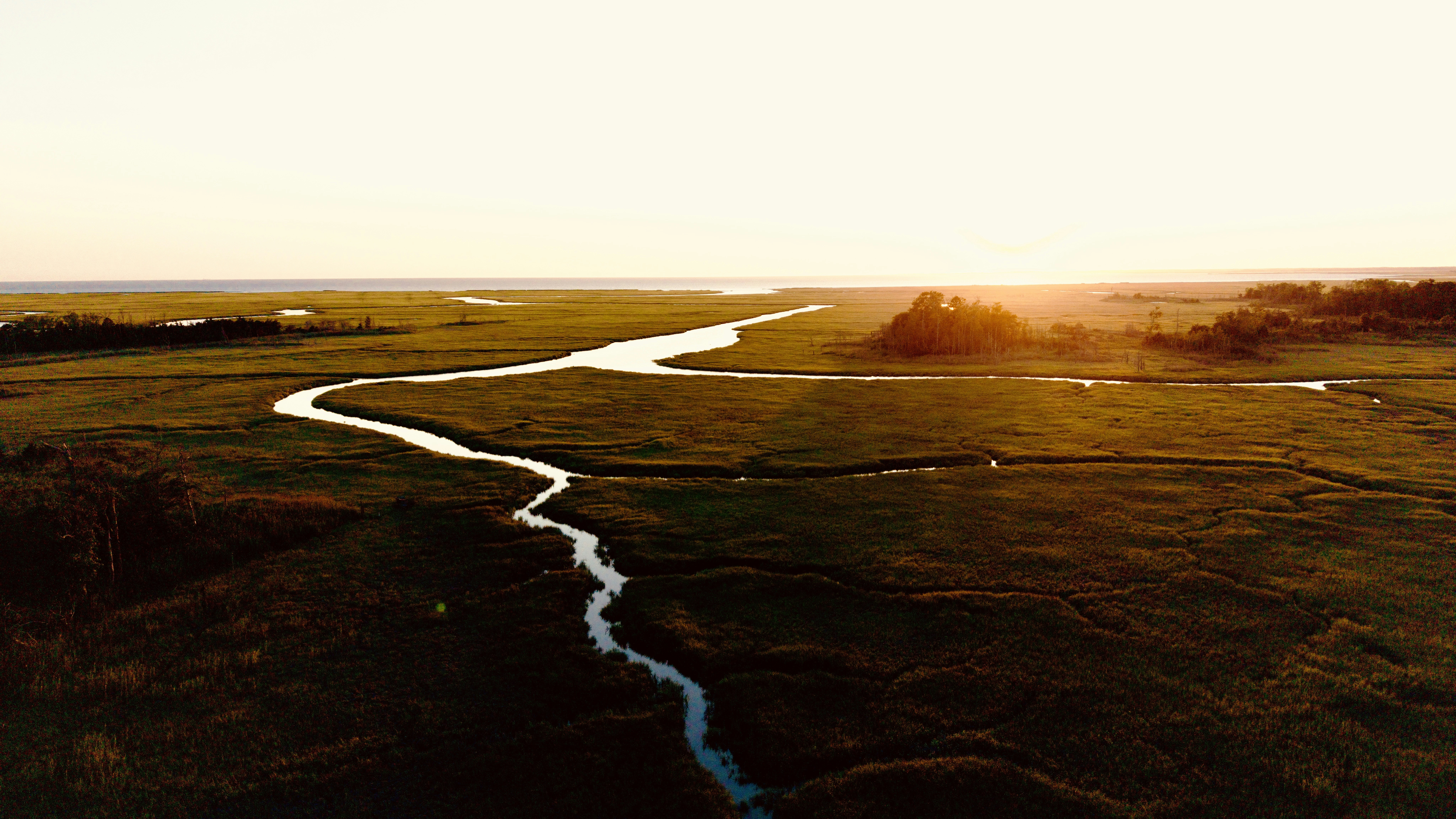 Dramatic coastal marsh with a meandering river catching the last light at sunset; a wide, low-angle view emphasizing endurance of the waterway and expansive sky. (Still image from landscape scene; no people or logos.)