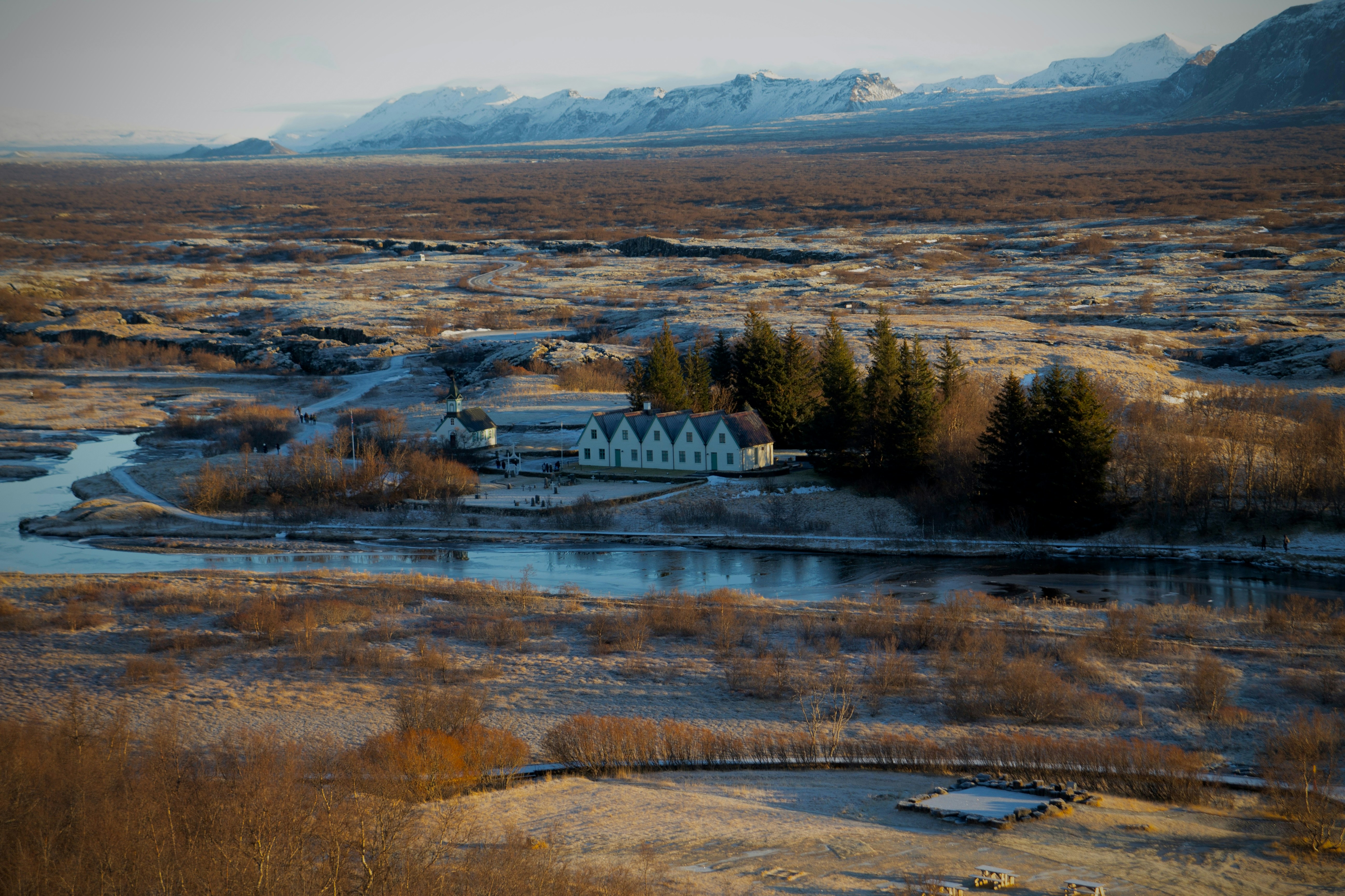 Low-lying farmhouse cluster sits beside icy marshes with distant snow-capped mountains under soft light at dusk. A tranquil, wide-shot landscape focusing on solitude and the small settlement amid expansive tundra.