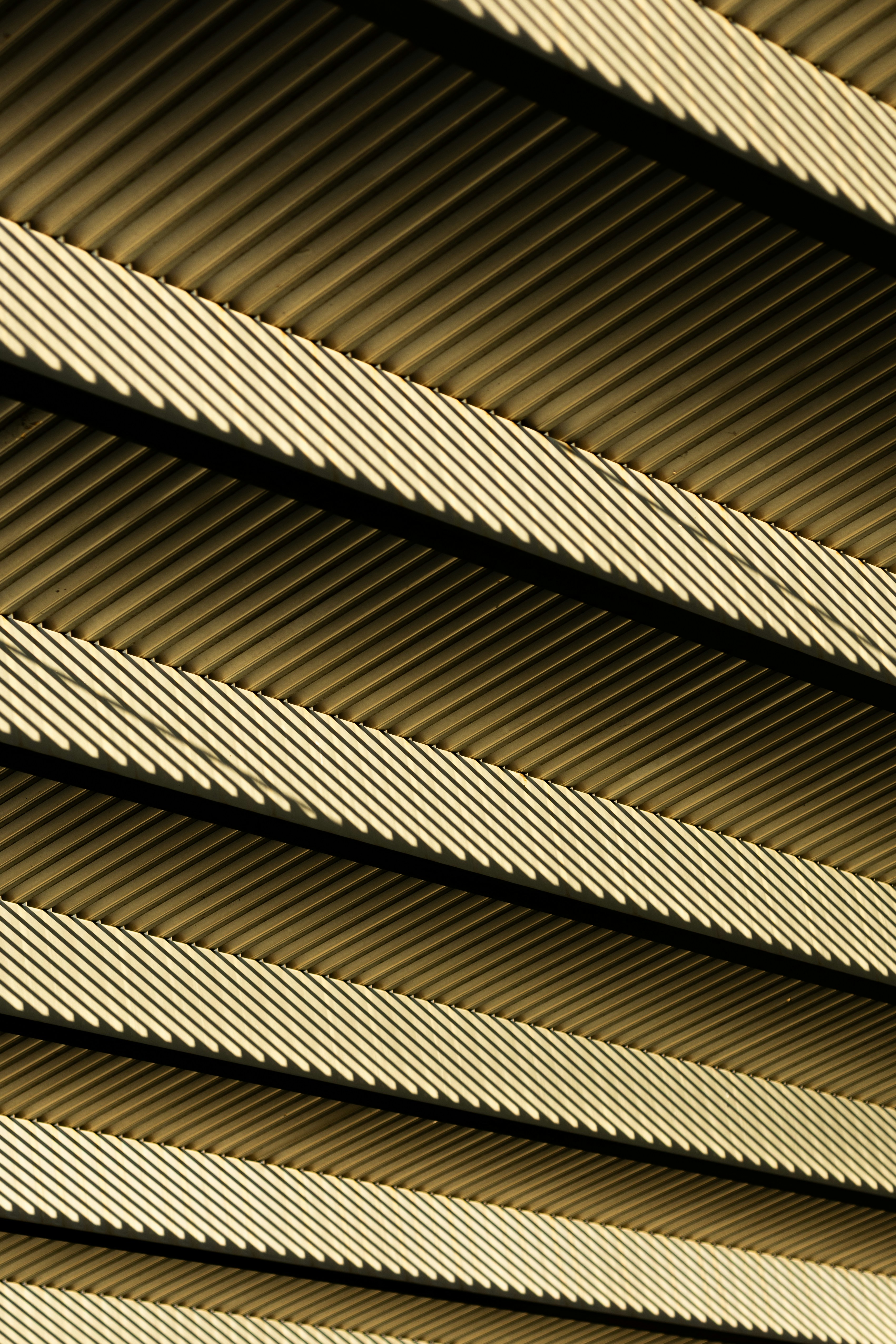 Golden diagonal louvers create a pattern of alternating light and shadow across the frame, emphasizing texture and repetition in a close-up architectural detail.