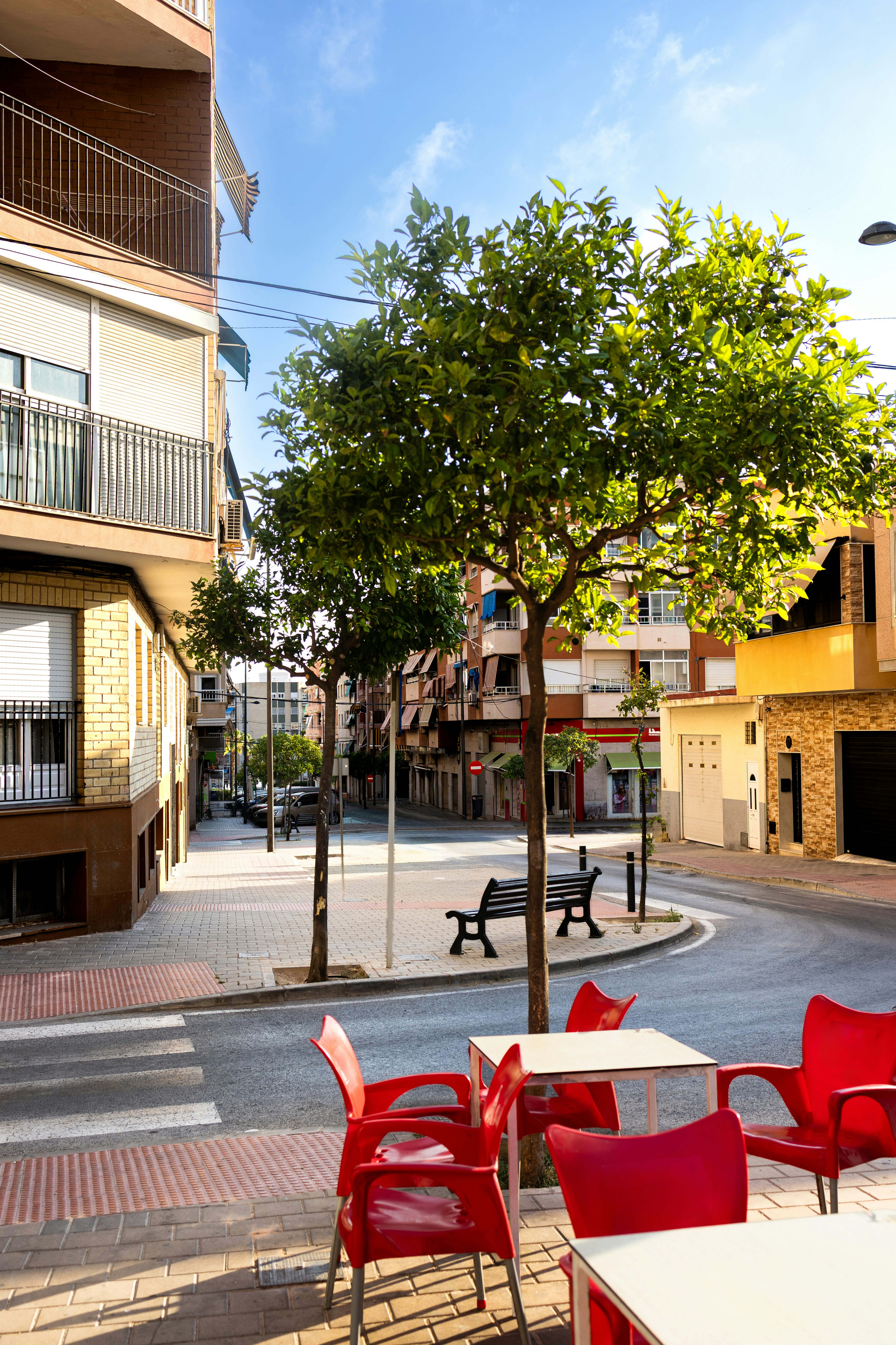 Sunlit Plaza: Trees, Benches, and Red Chairs in a Quiet Street