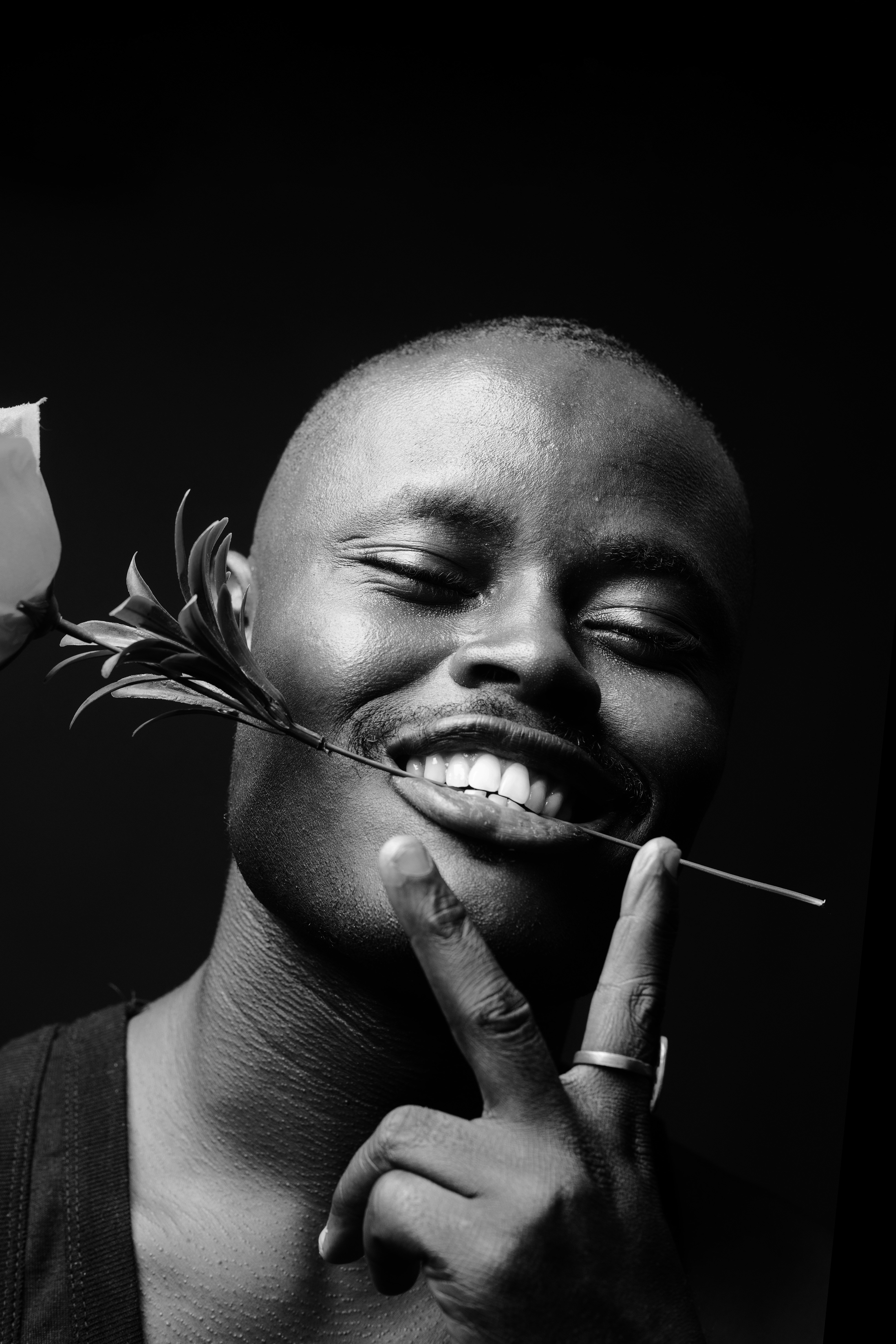A smiling man holds a flower near his mouth, the stem gently crossing his lips; the shot emphasizes the playful pose and relaxed expression. This is a black-and-white portrait.