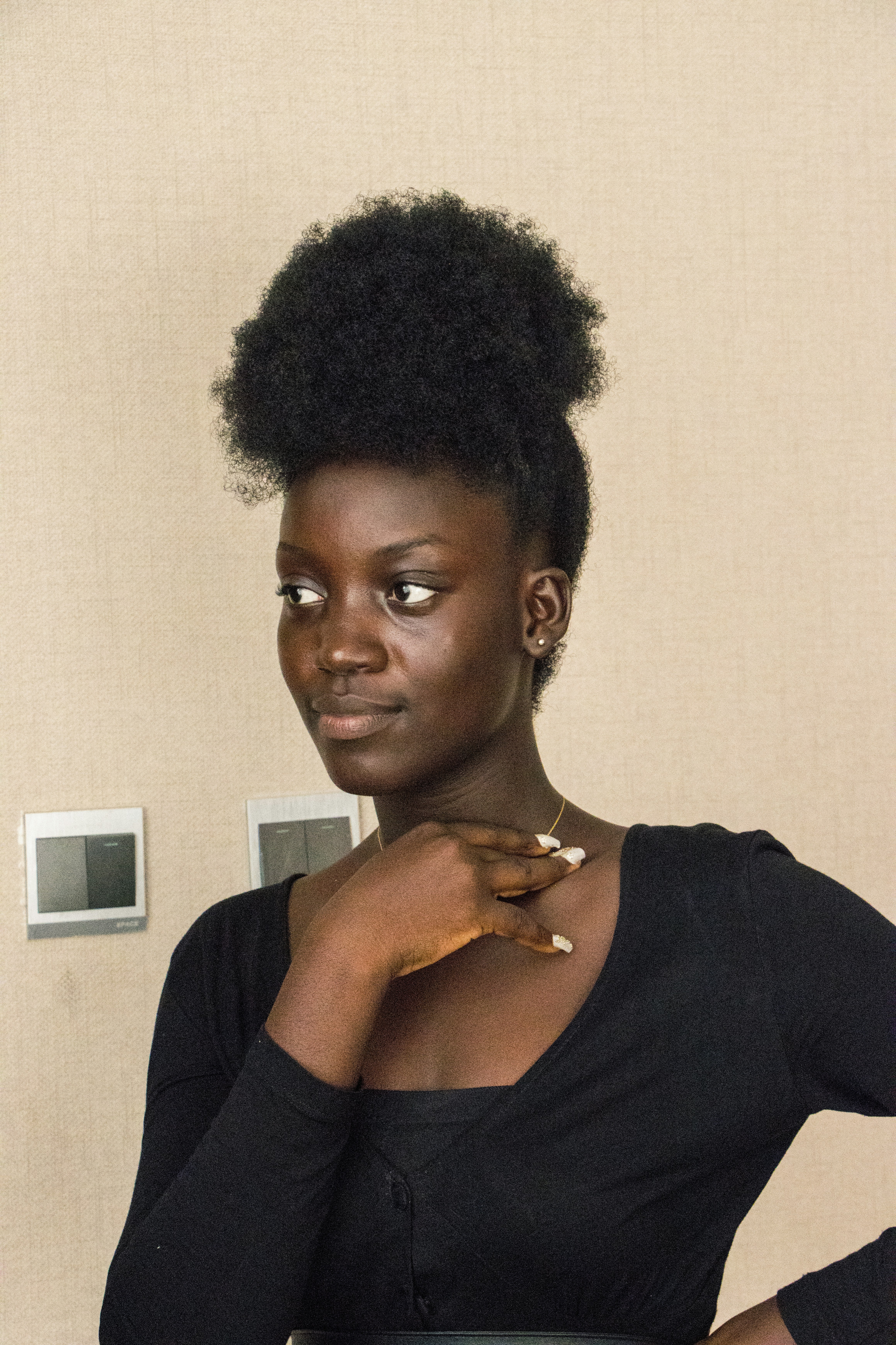 Portrait of a thoughtful woman with natural hair updo, posed against a neutral wall; the composition centers her face and hand near the collarbone, highlighting calm expression. Neutral-toned background suggests an indoor studio setting.