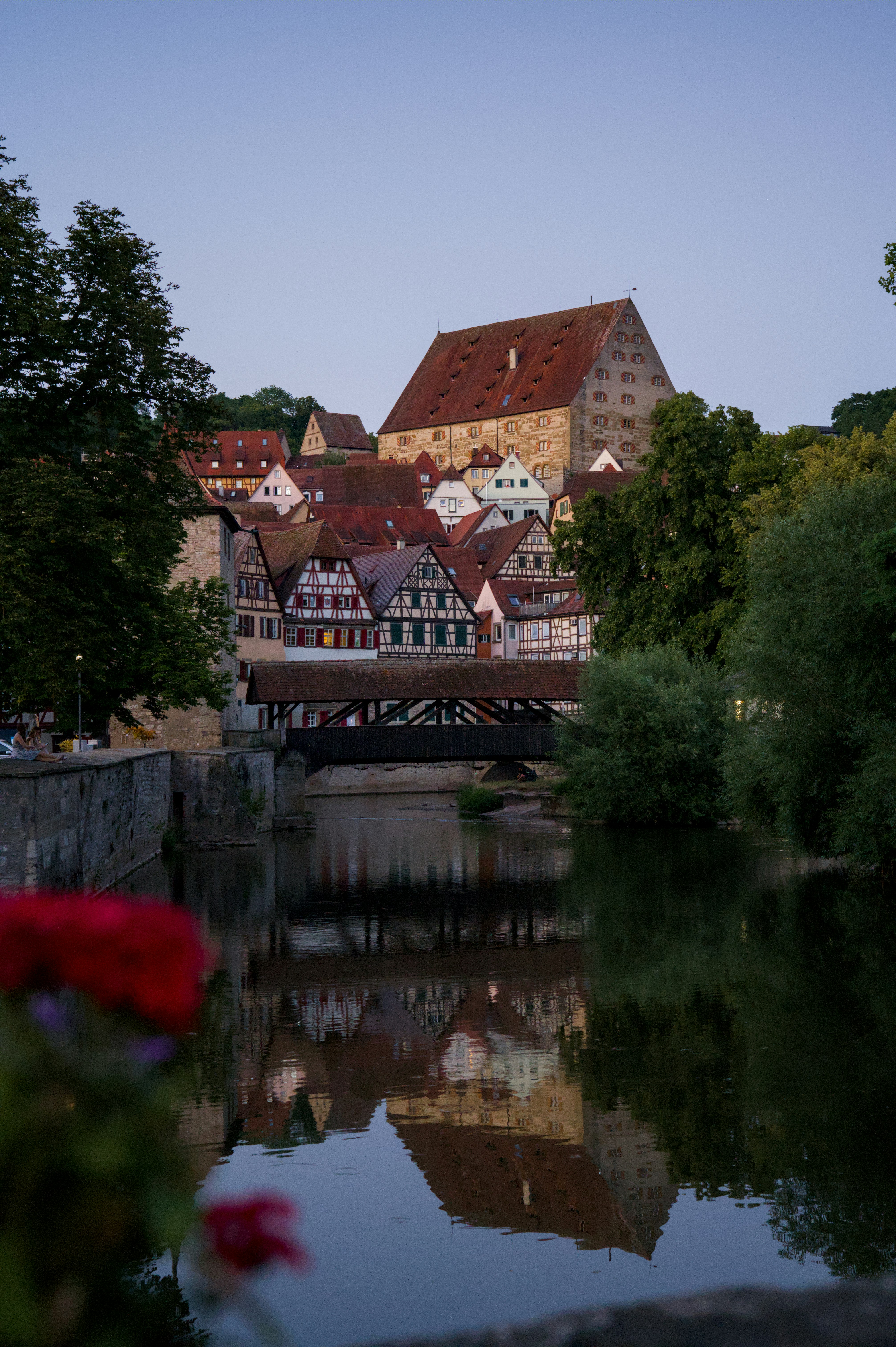 Historic town with timber-framed houses along a calm river, a stone bridge, and a large old building on a hill; foreground flowers and trees frame the scene.